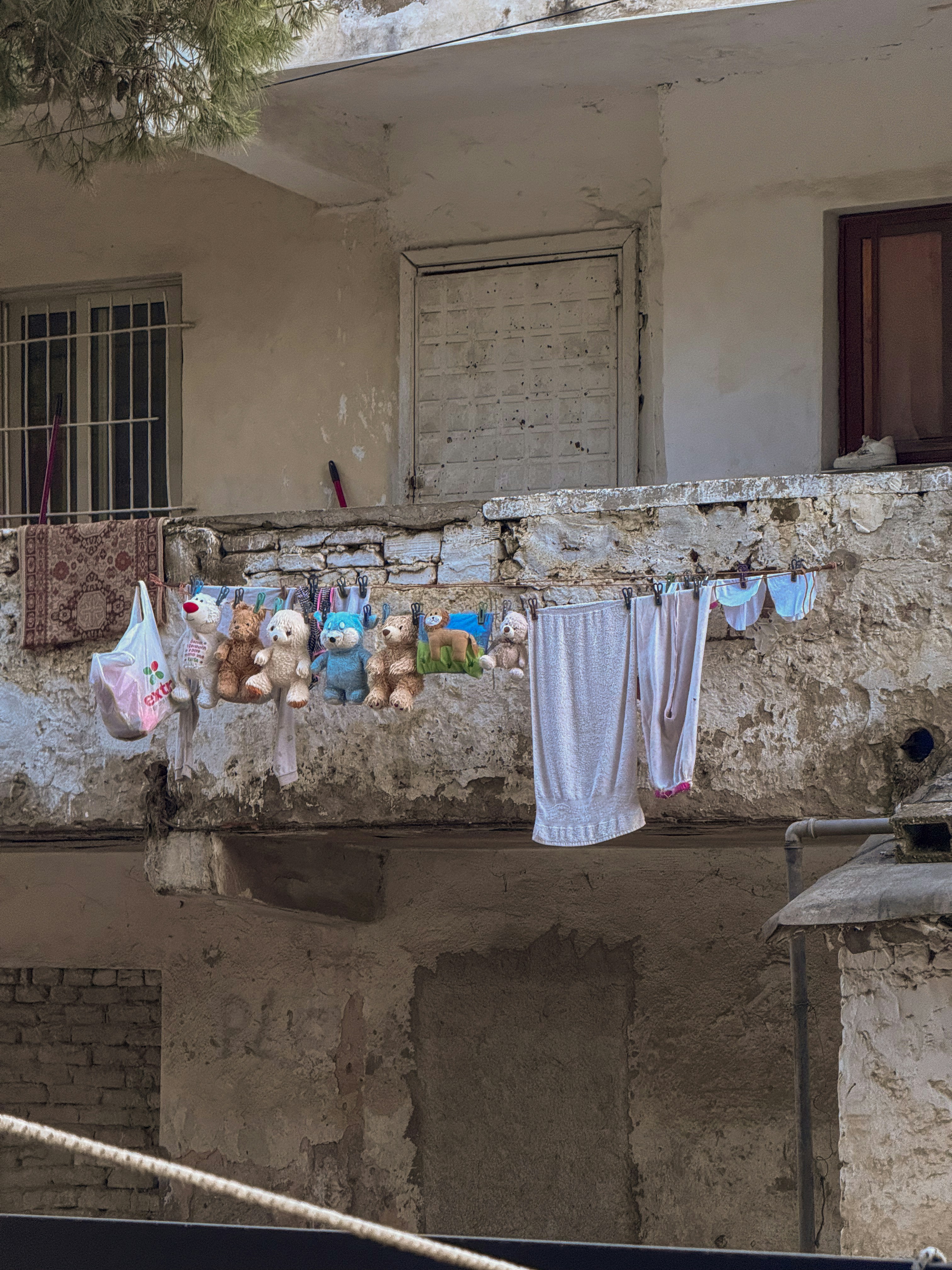 Rows of stuffed animals and clothing lines hang between weathered walls on a sunlit balcony, capturing a quiet domestic scene with a sense of resilience.