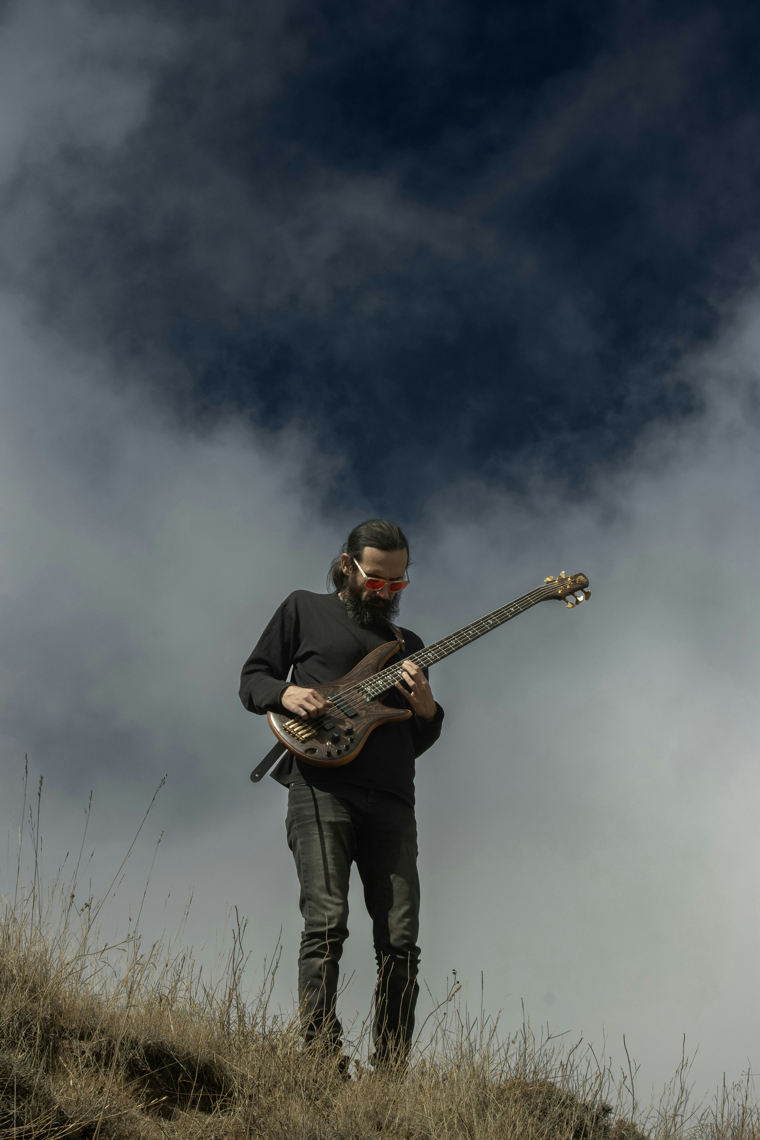 A man with a beard playing a bass guitar on a grassy hill