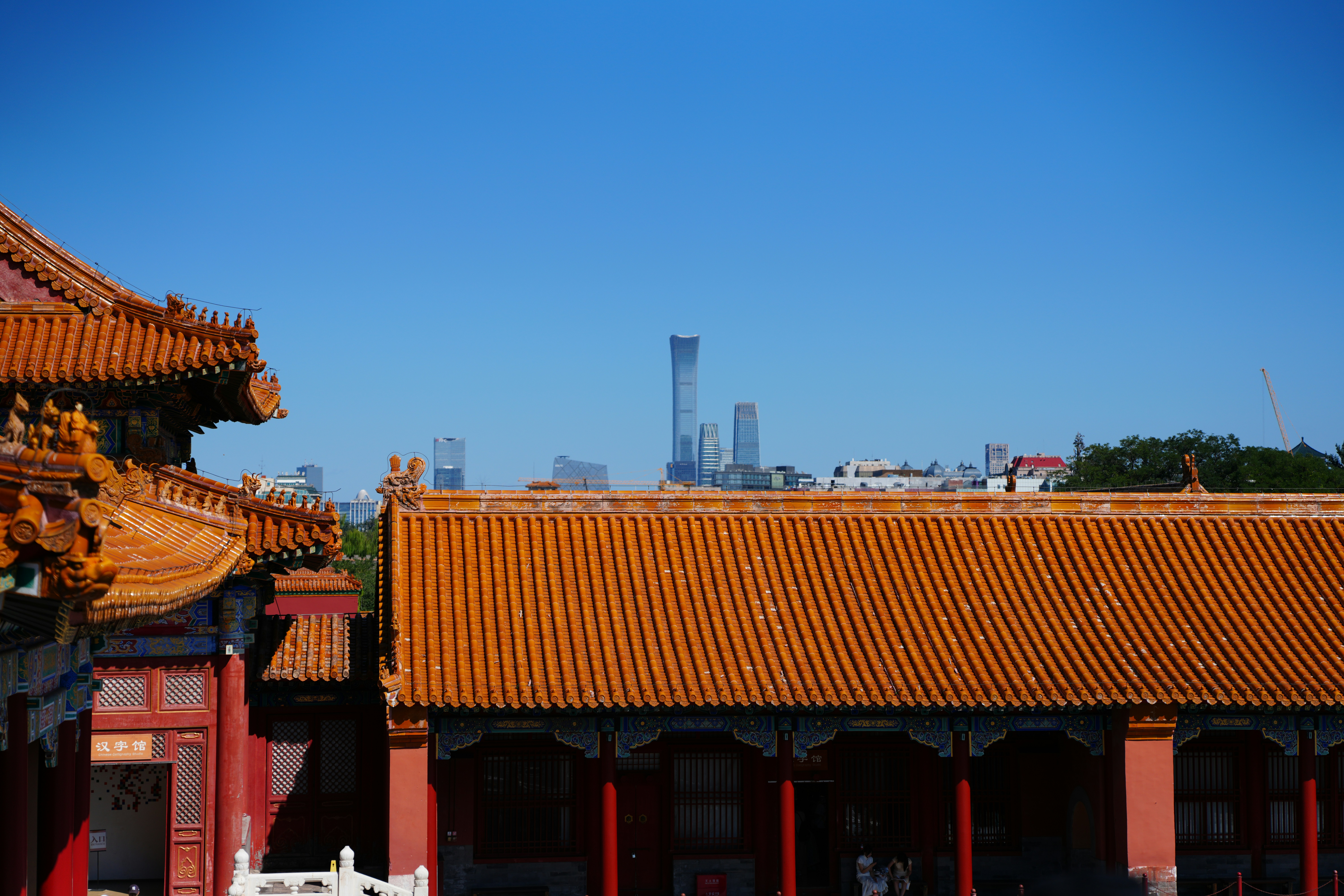 Red-tiled temple rooftops frame a bright blue sky with distant high-rise silhouettes in the background. A traditional architectural foreground contrasts with the urban skyline beyond.
