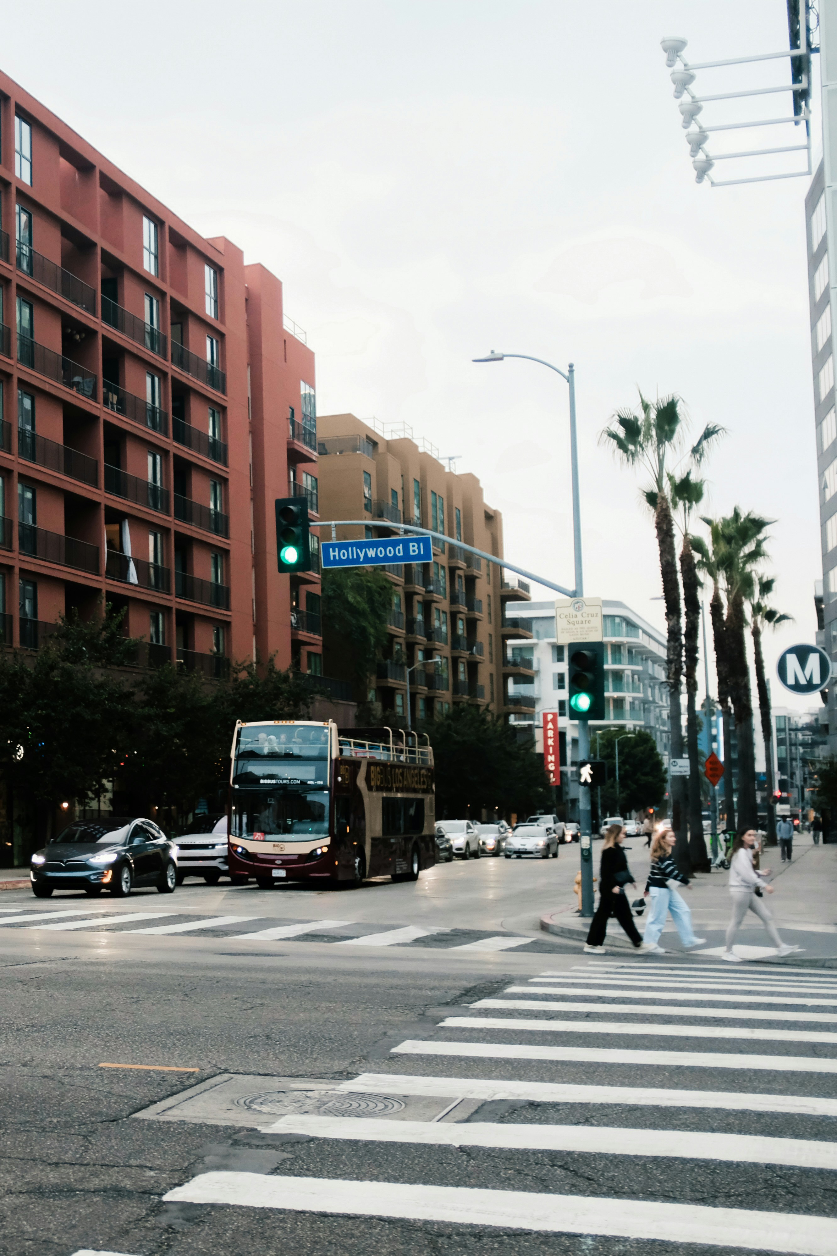 Busy city intersection with pedestrians crossing a wide crosswalk as a bus and cars navigate a tree-lined street near colorful apartment blocks.