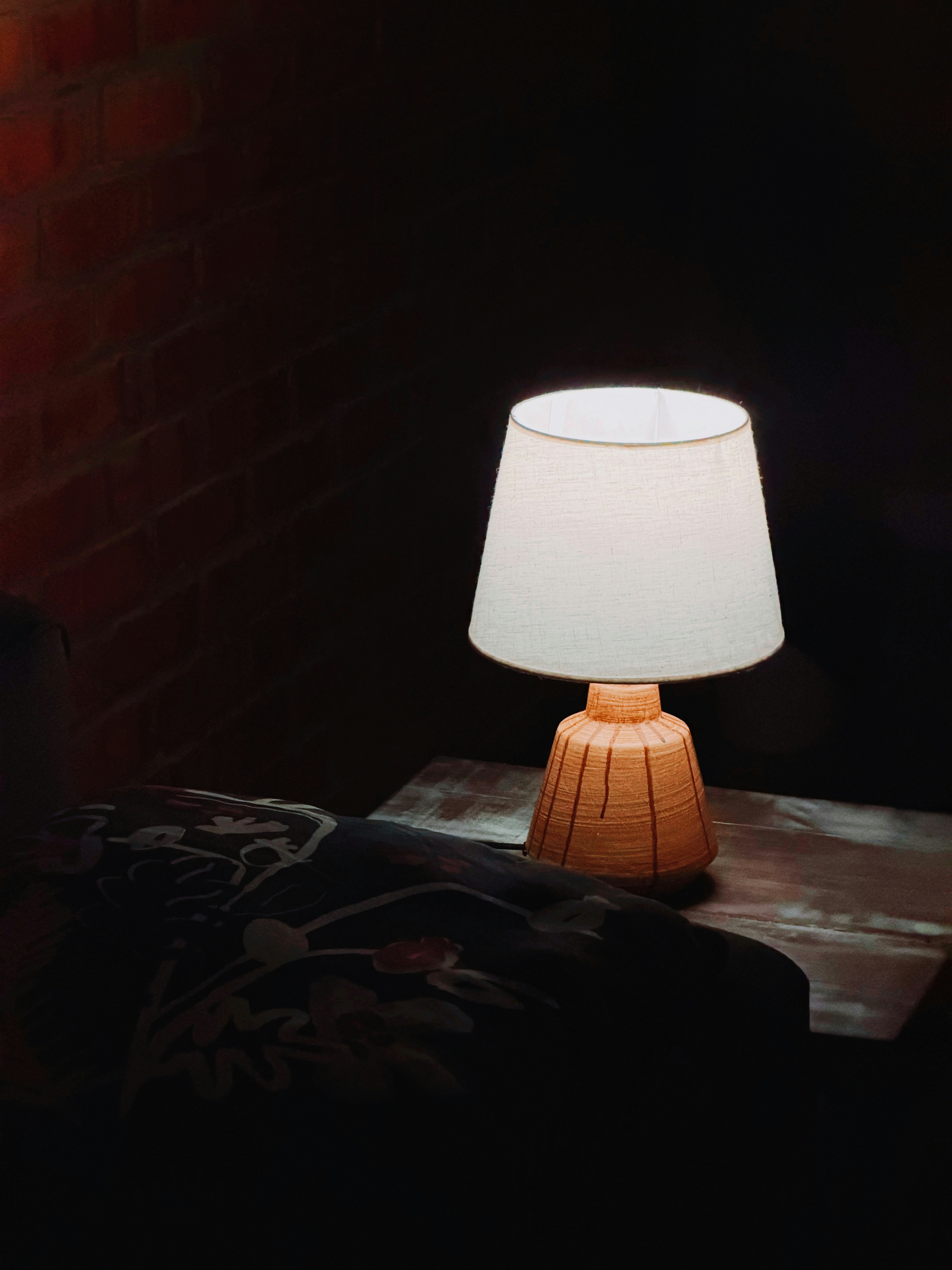 A bedside lamp on a wooden table emits a warm glow in a dim room, with a textile item partially visible in the foreground. The scene emphasizes cozy lighting and intimate atmosphere; a small logo-like lamp sits as the focal point.