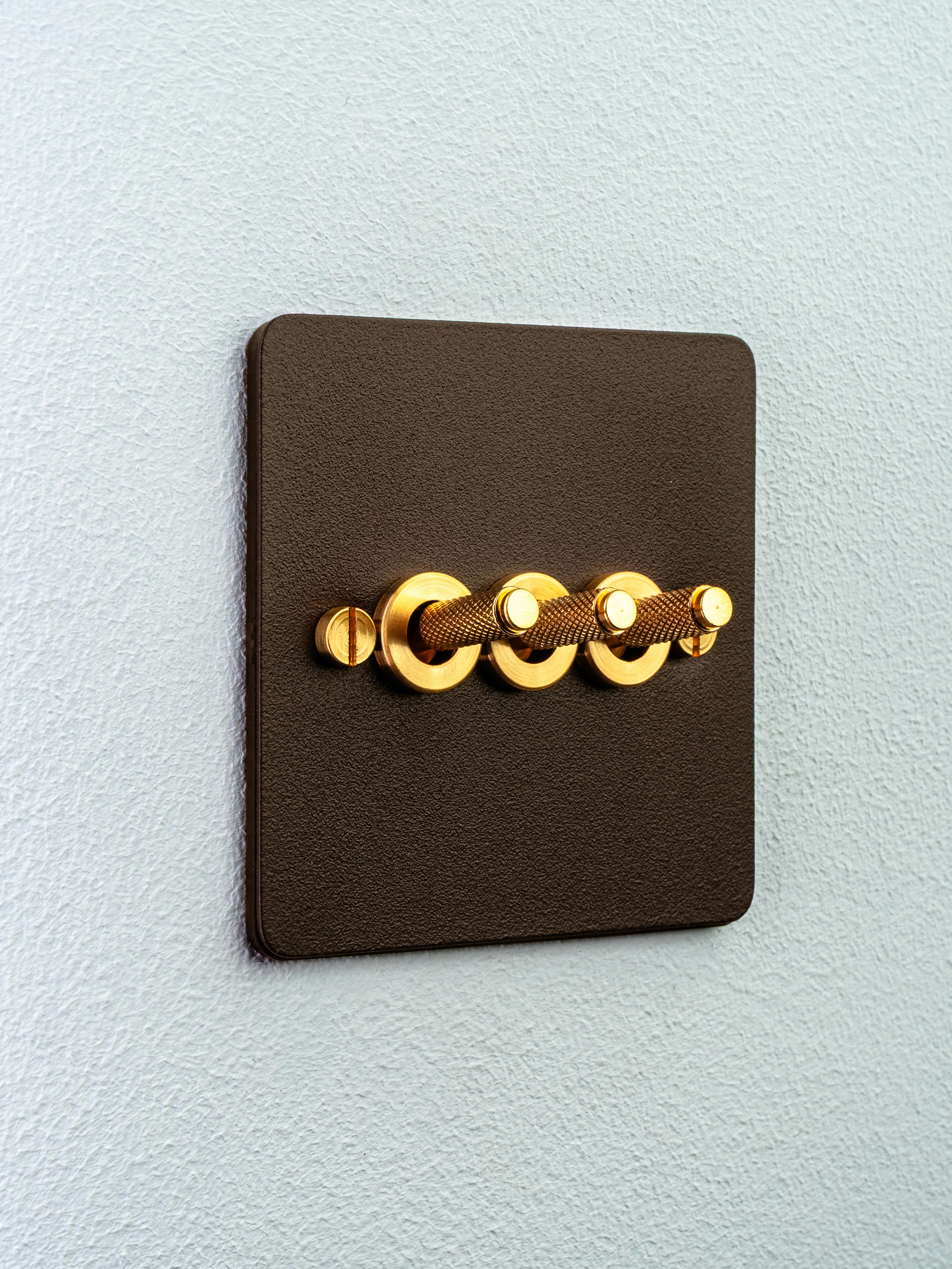 Close-up of a metallic gold coat hook rail mounted on a dark square backing, set against a pale textured wall.
