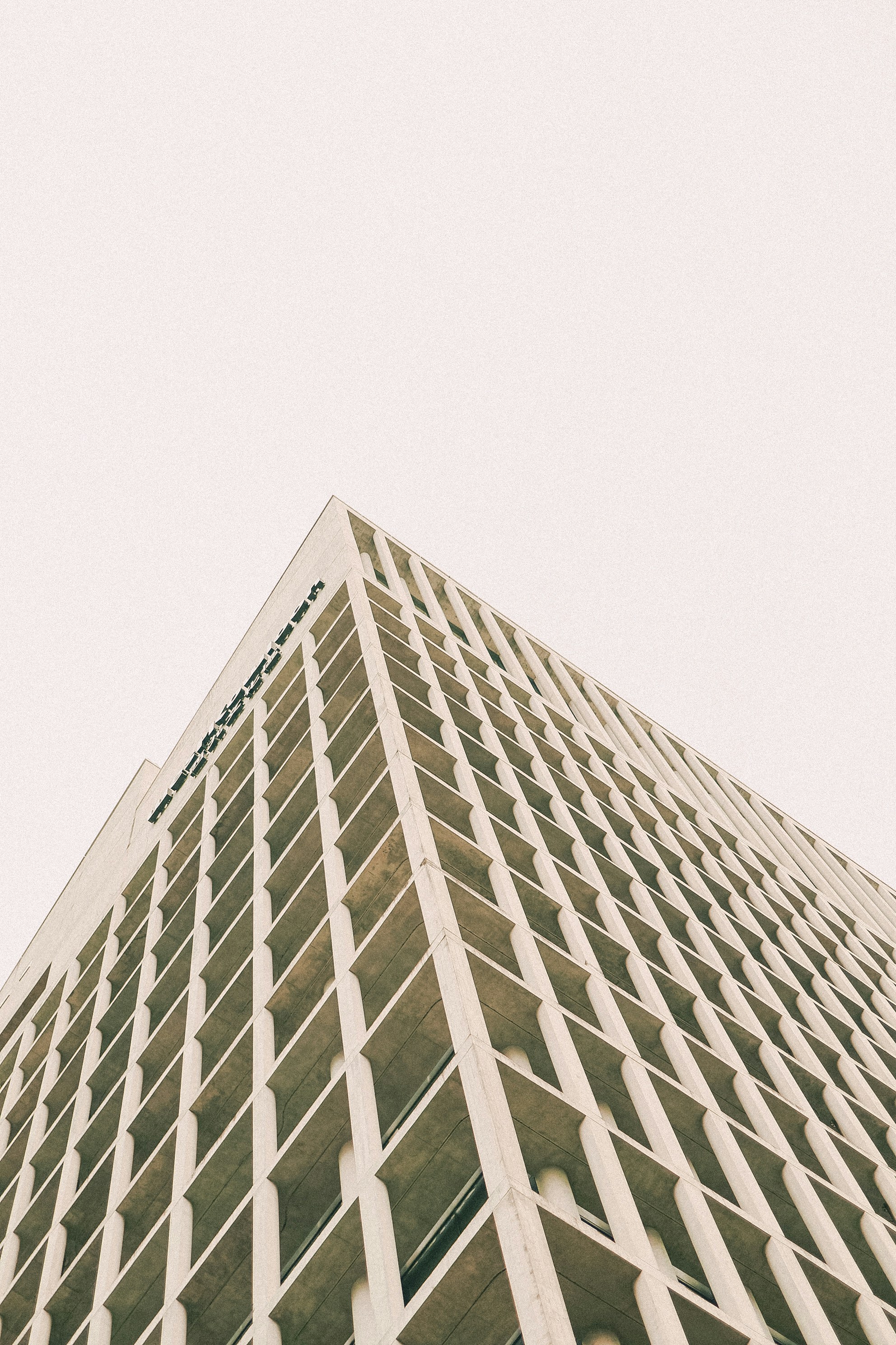 Concrete building with a repetitive grid pattern tilting upward, photographed from a low angle; minimal color palette with soft, pale sky in the background.