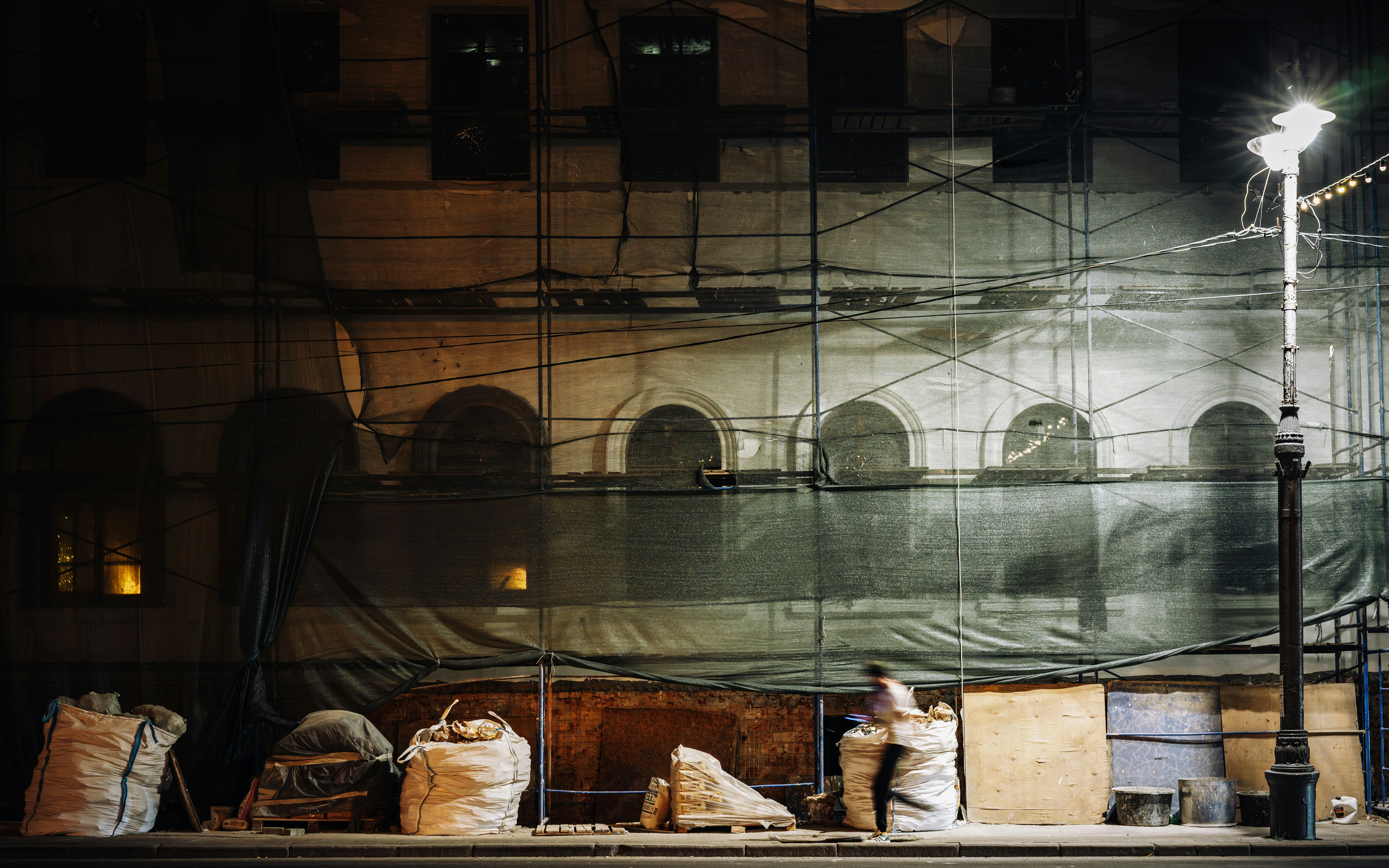 Dimly lit alley with bags and crates stacked along a street under a pale urban glow; a lone figure trails across the scene, suggesting a city repair or construction work at night.