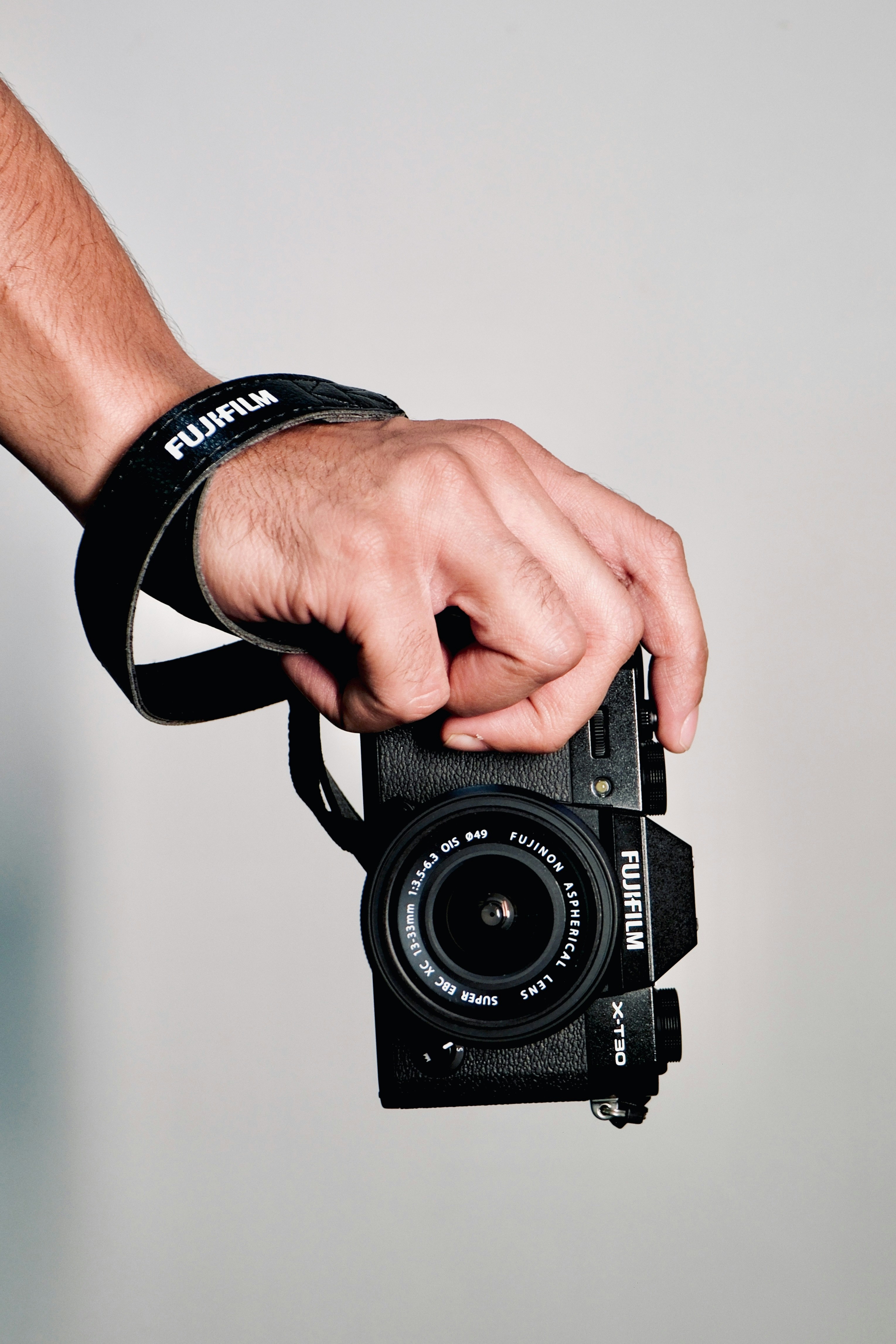 A close-up of a hand gripping a compact Fujifilm camera against a neutral backdrop; the device's lens and branding are clearly visible. This is a product or lifestyle shot featuring a real camera as the subject.