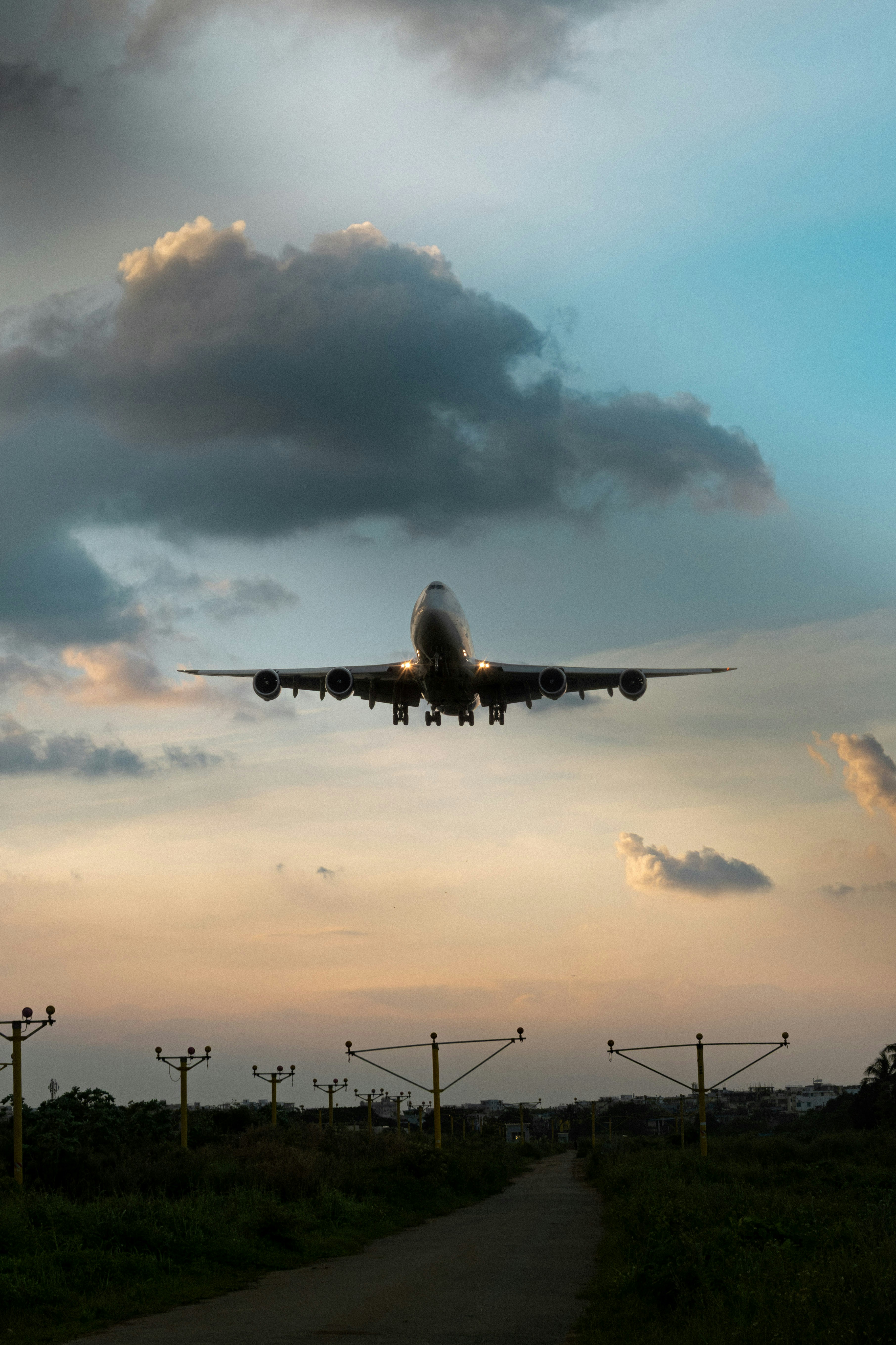 Silhouette of a large commercial aircraft on final approach above a rural runway at sunset; low foreground reveals a path and equipment, with dramatic clouds and warm sky coloring. (Photo is a photographic capture, not a drawing or painting.)