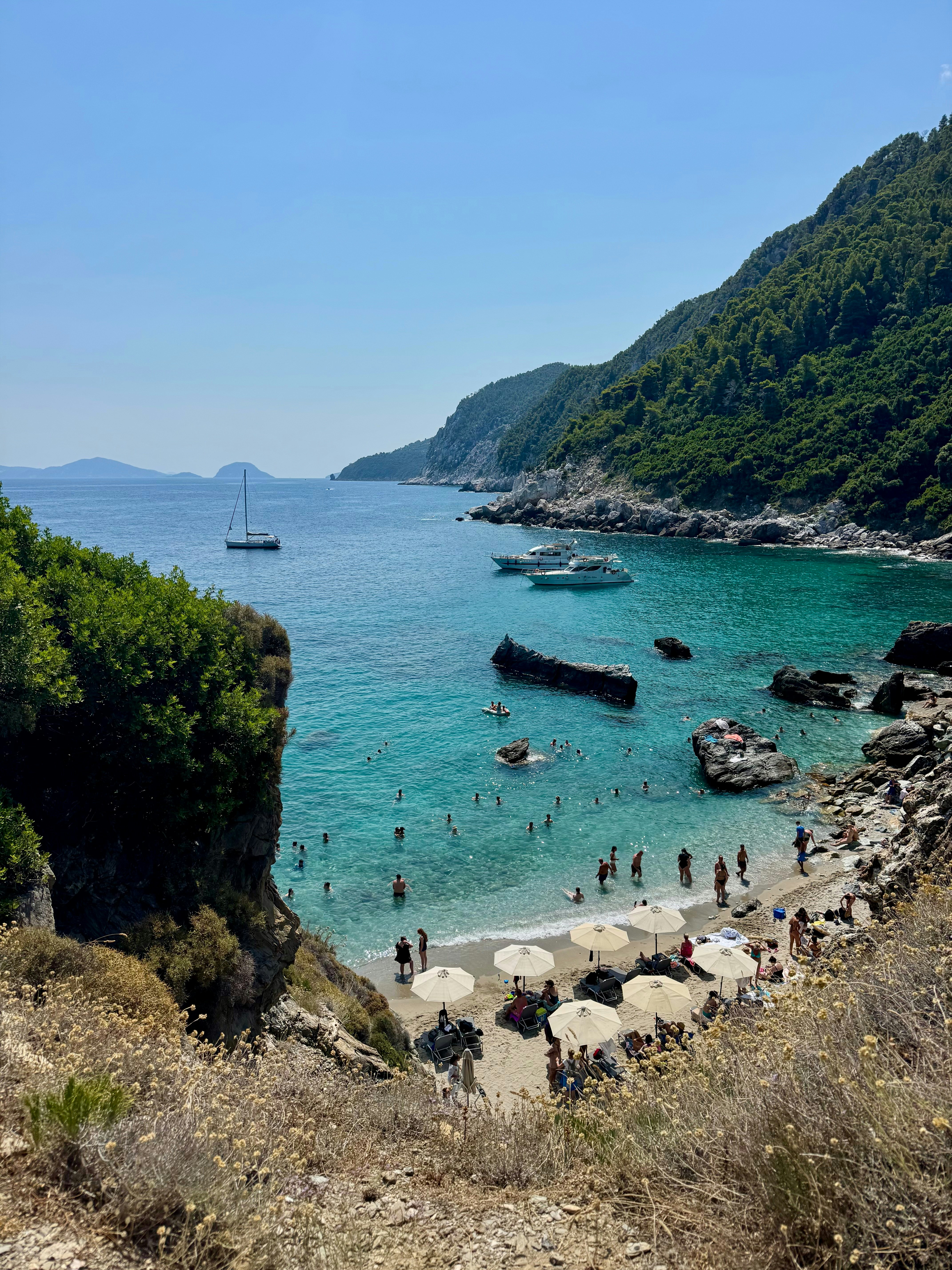 Bustling beach cove with turquoise water, sunbathers under umbrellas, anchored boats, and rocky cliffs along a clear coastal inlet.