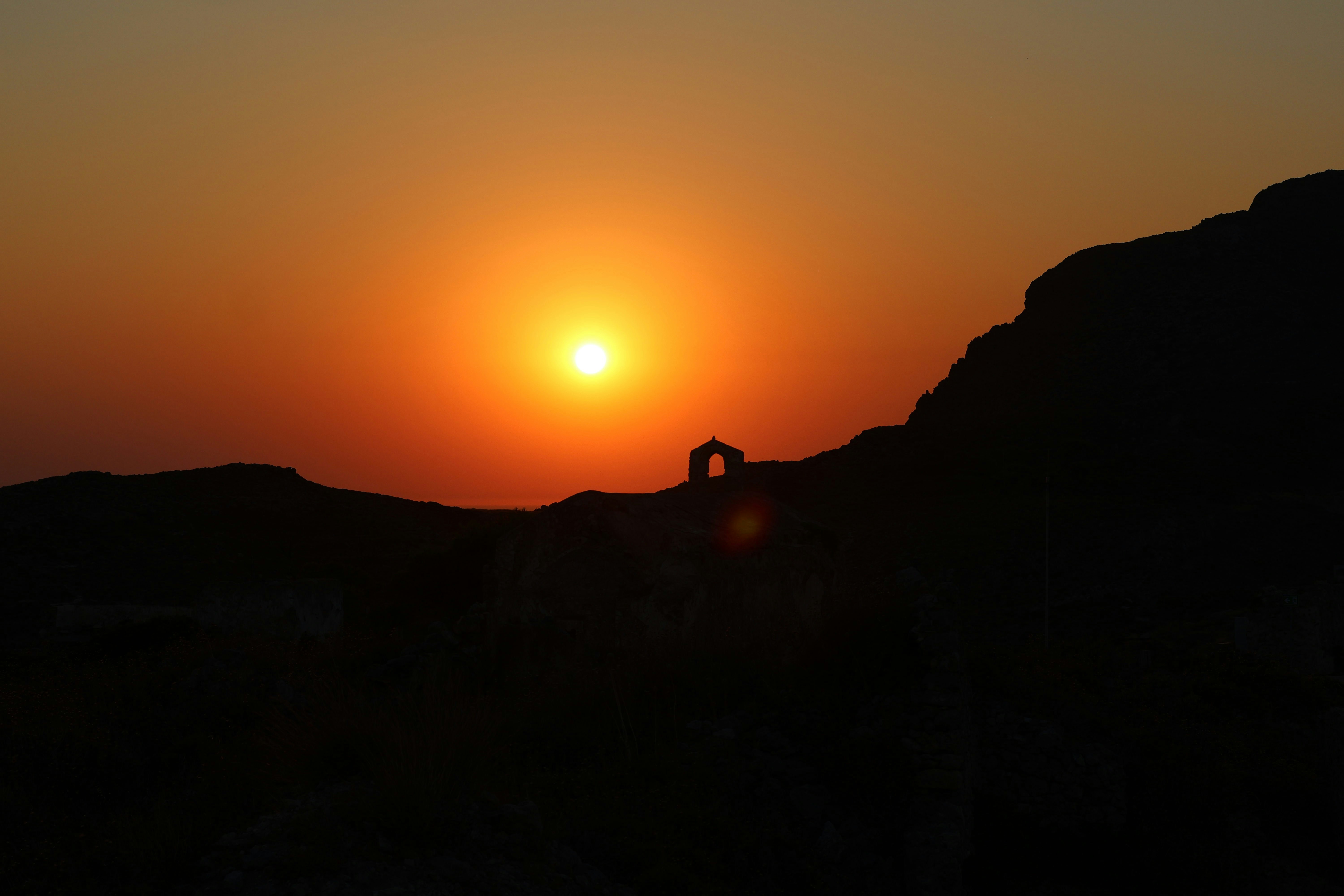 Low-sun silhouette of a rugged ridge with a small arch-like structure near the horizon; warm gradient sky and minimal foreground detail.