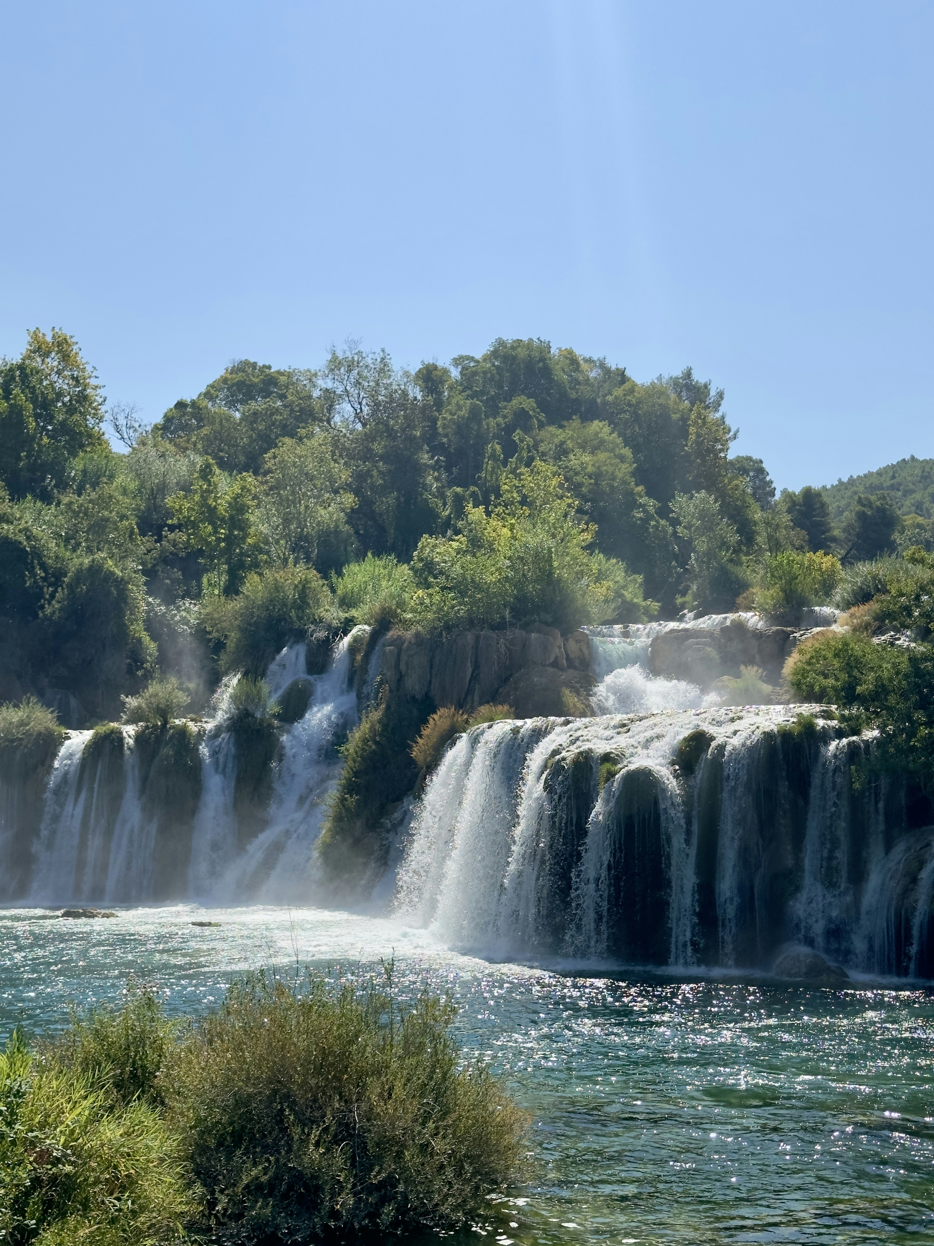 Vibrant waterfall cascading over rocky ledges with lush green trees and a bright blue sky in sunlight.