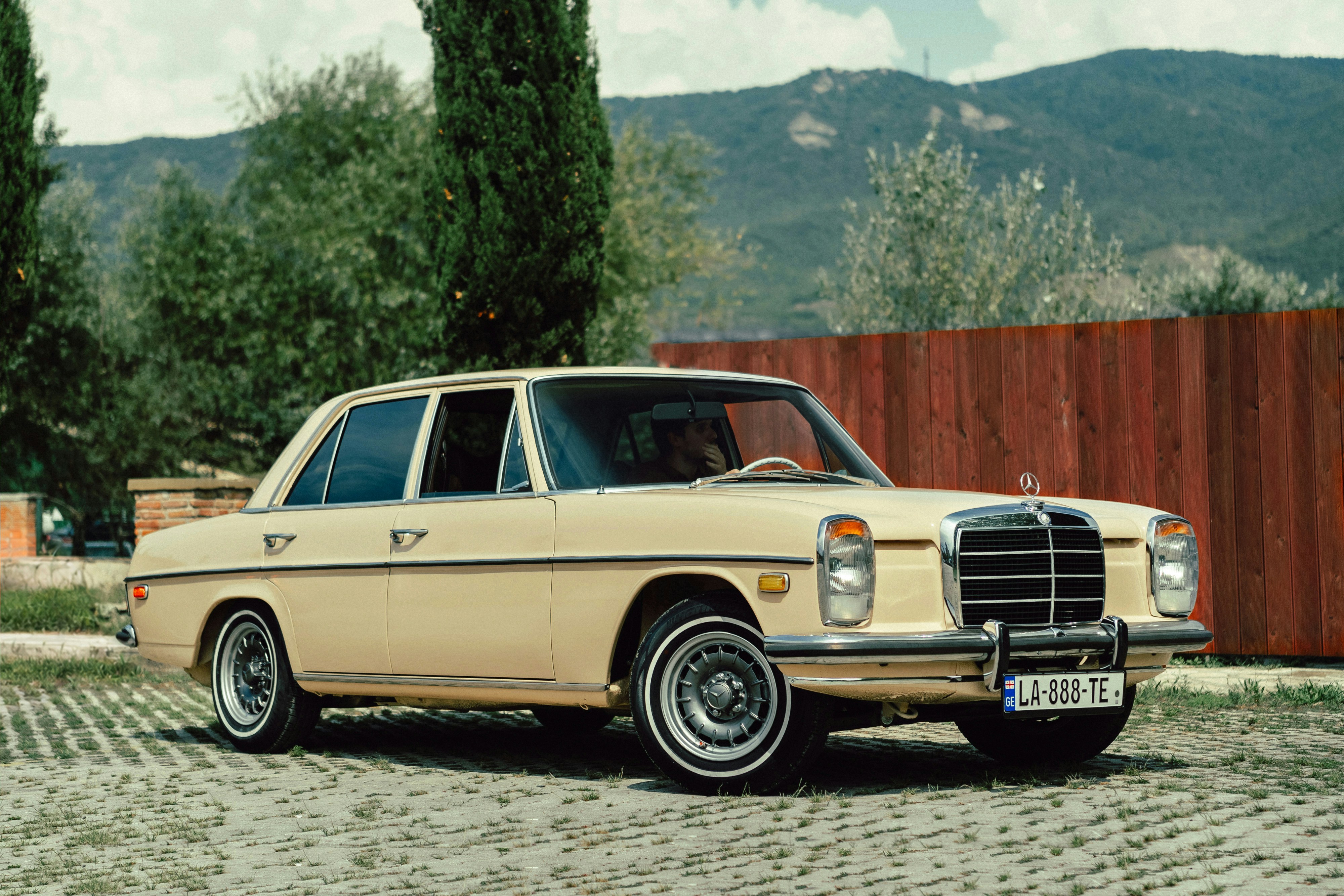 Cream-colored classic Mercedes on a cobblestone lot with a red wooden fence and distant hills in the background.