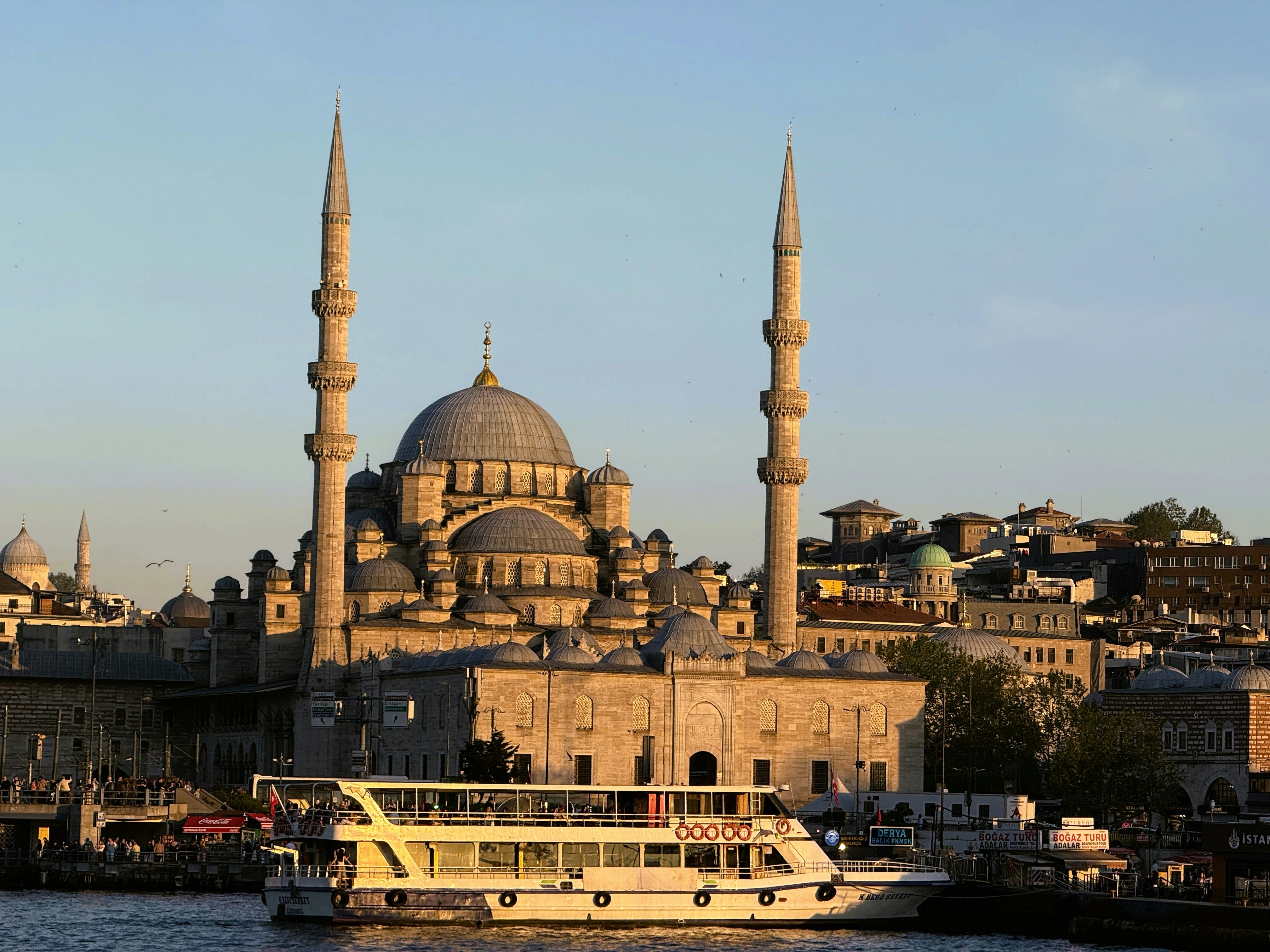 Dome and minarets of a historic mosque rise above a riverside district with a double-decker boat in the foreground.
