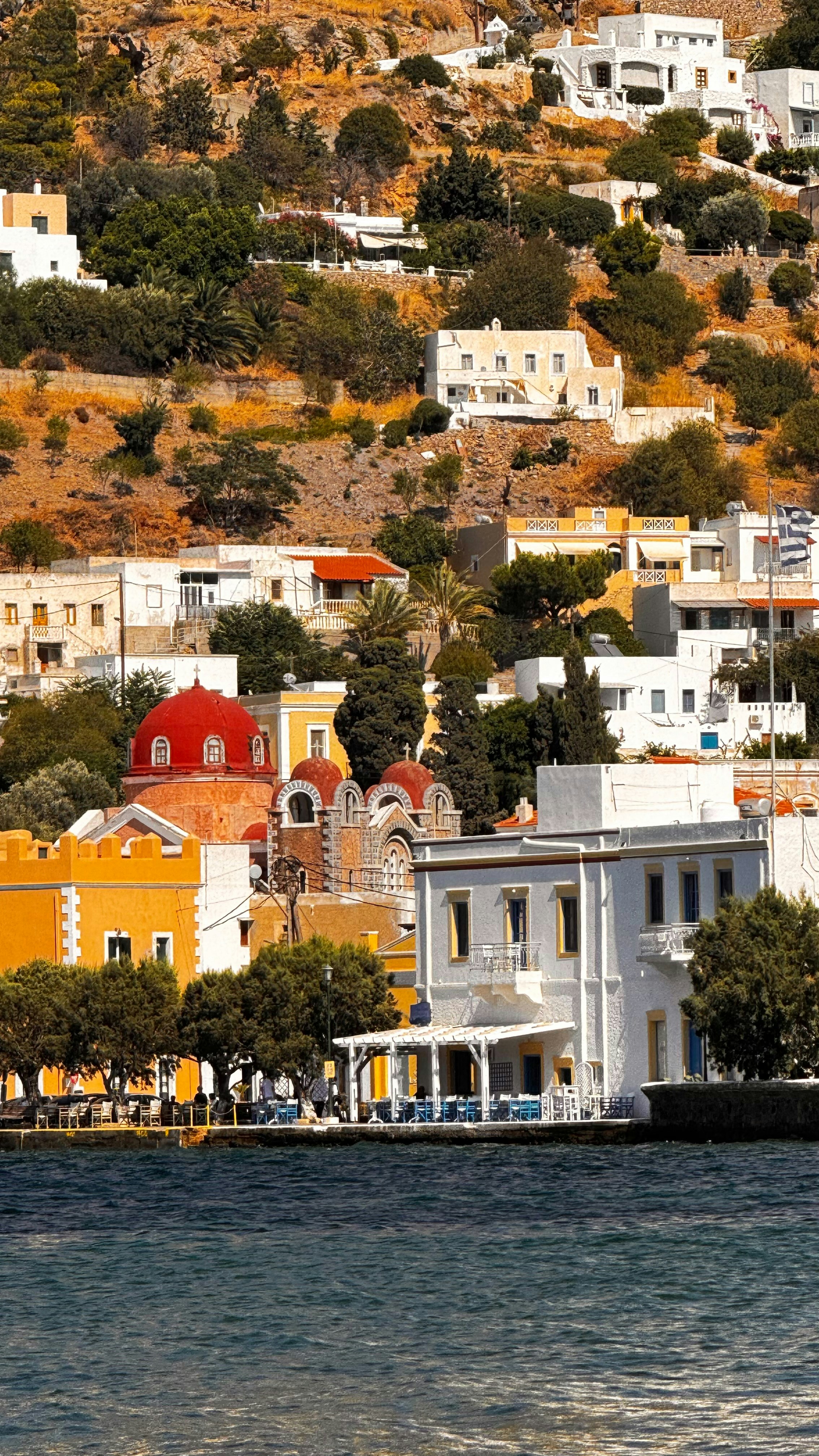 Houses cling to a sunlit hillside above a tranquil harbor, with a colorful Mediterranean palette and a red-domed structure near the waterline.