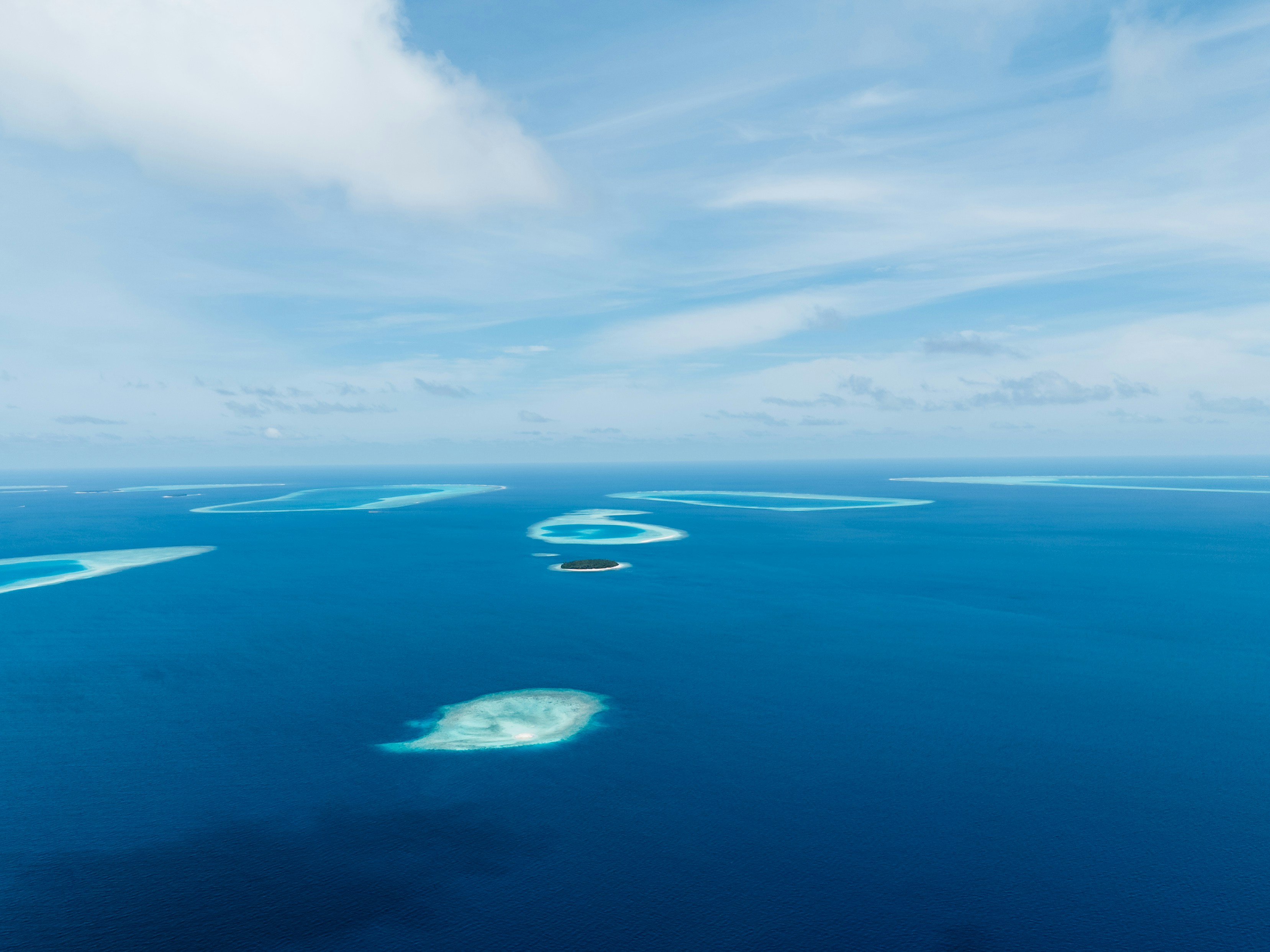 Aerial view of bright turquoise lagoons and pale sandbars forming winding loops across a deep blue sea, seen from high altitude. The image captures a landscape of shallow atolls and small islets beneath a soft cloud-filled sky.