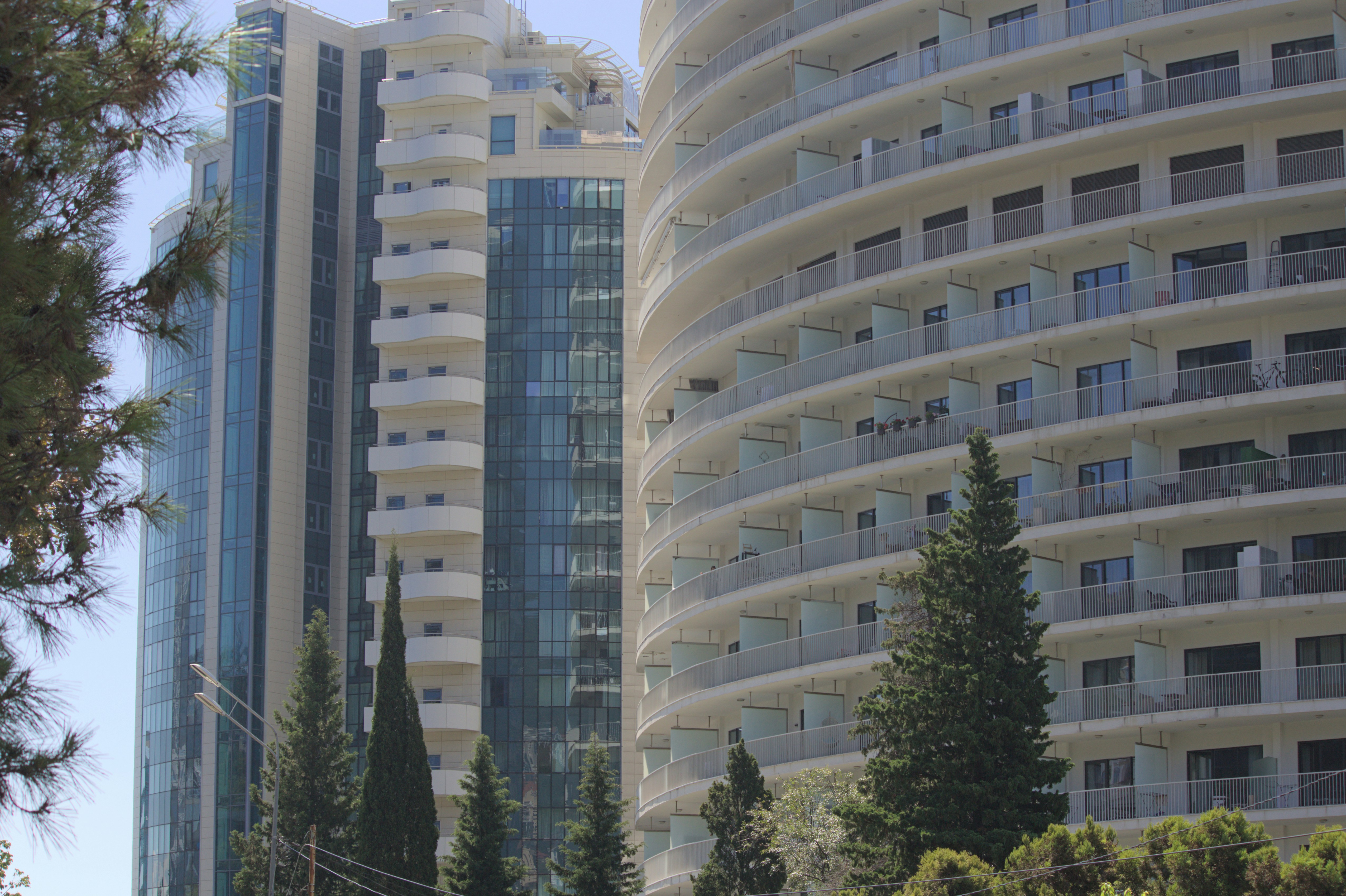 Two modern high-rise buildings with curved balconies rise over a landscaped foreground of trees and evergreens.