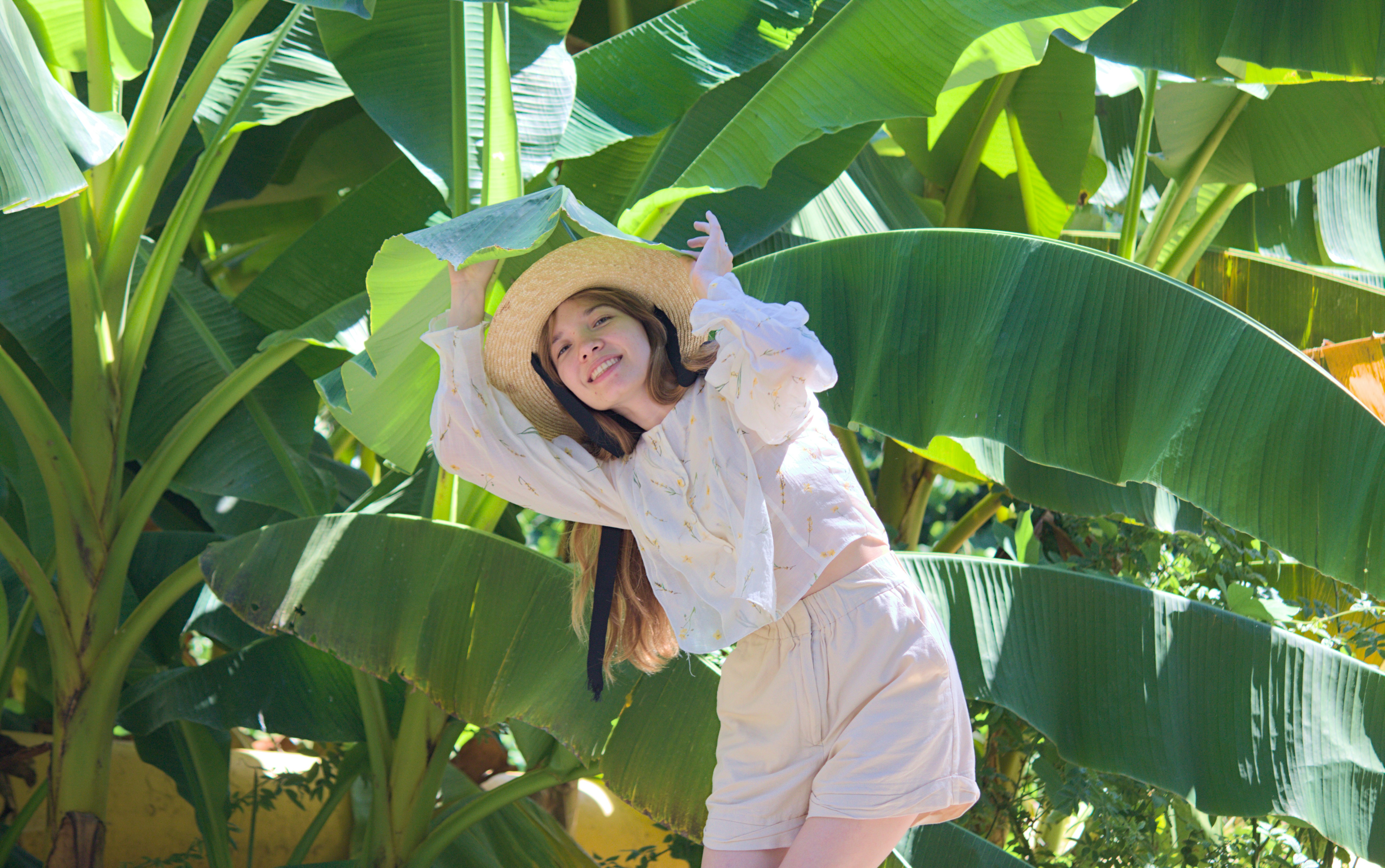 A smiling young woman in a straw hat and white blouse playing hide-and-seek under a giant banana leaf in a tropical garden.
