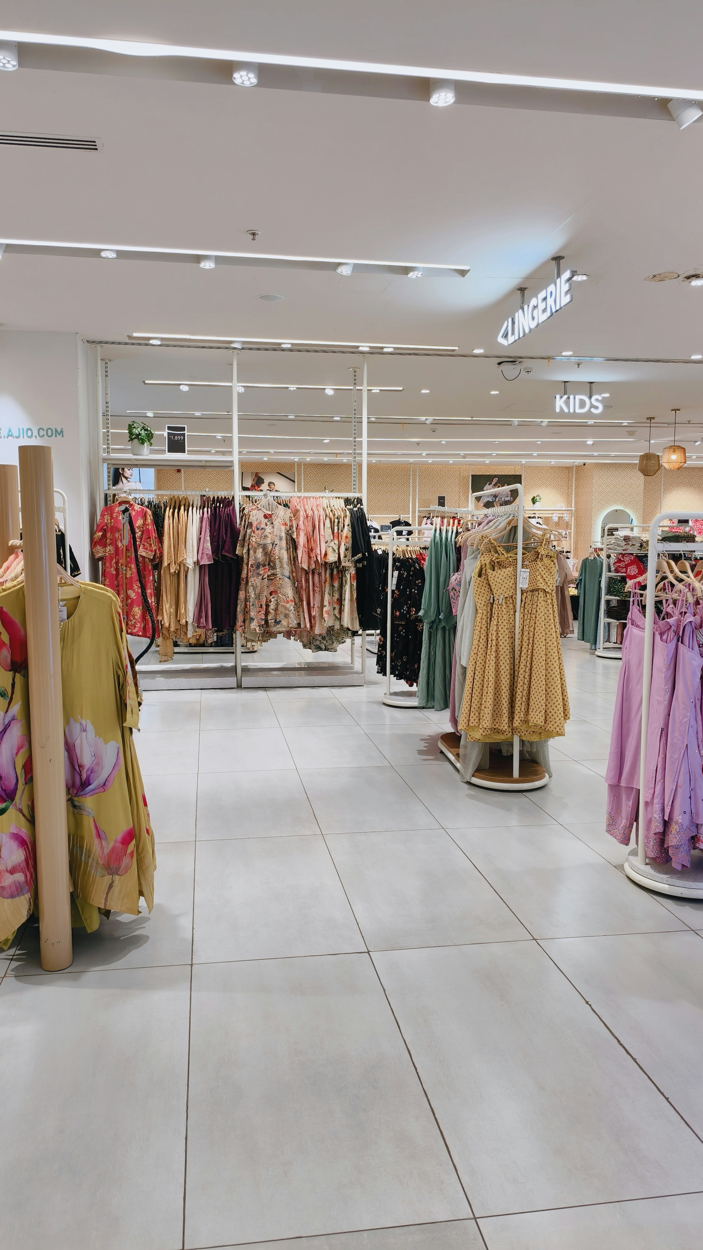 Brightly lit clothing store with racks of colorful dresses arranged along a wide aisle; signage for kids and lingerie visible in the background.