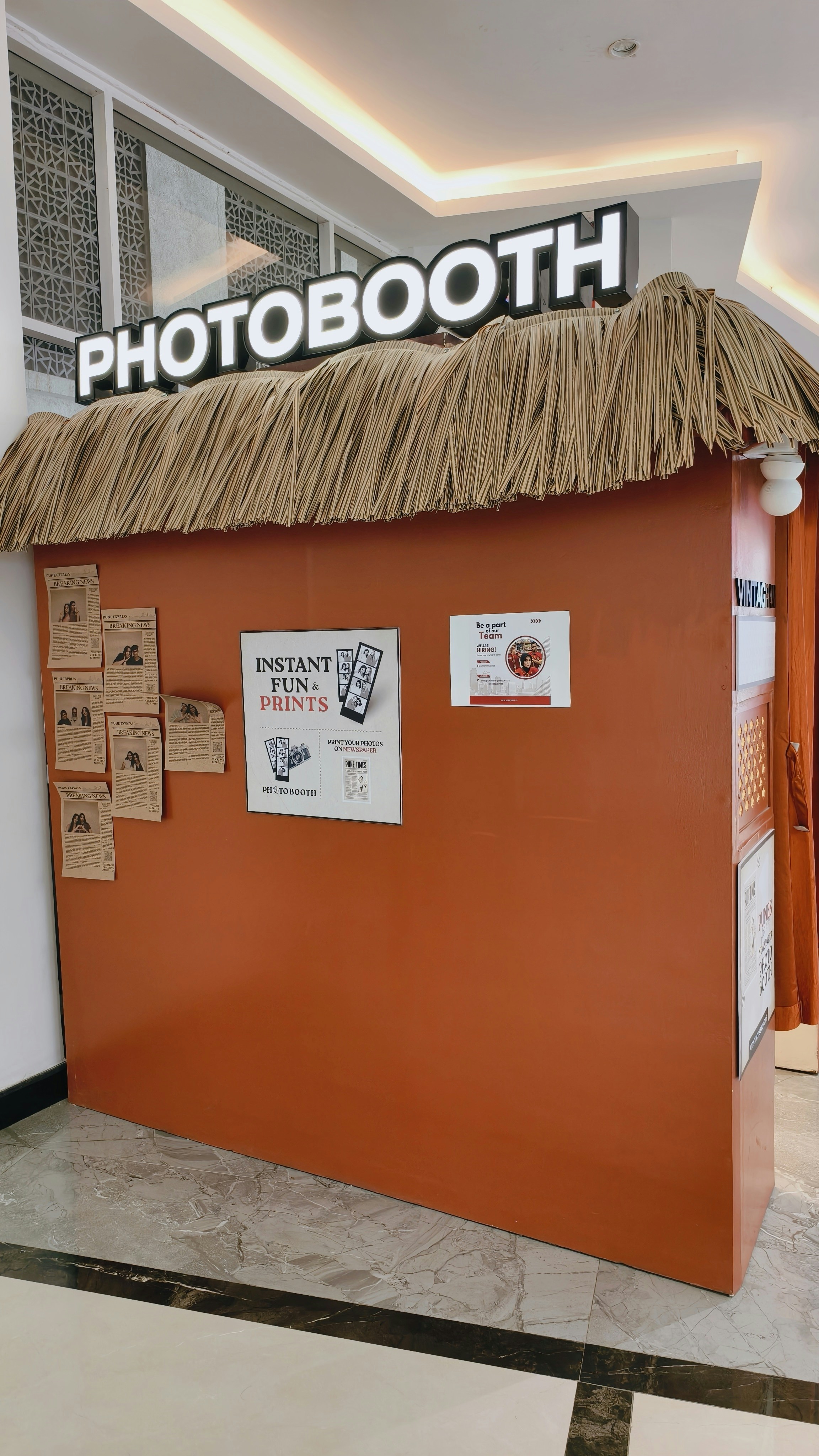 Vibrant orange photobooth booth with a thatched roof and bold white sign; scattered posters adorn the wall for a playful, retro vibe. This is a built setup inside a mall or lounge area.
