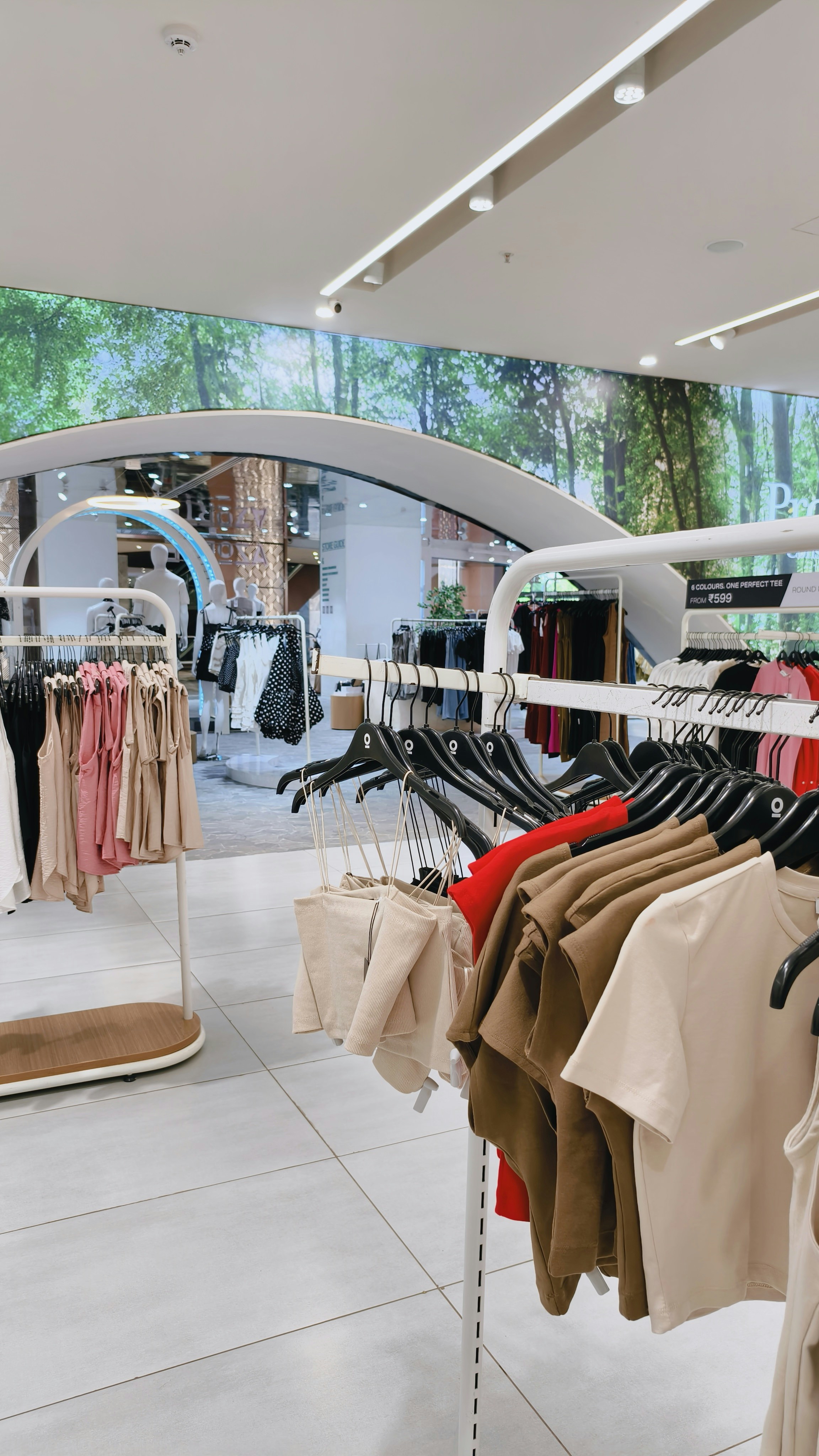 Retail clothing display in a bright department store with racks of shirts and dresses, foregrounded by curved white fixtures and a nature-themed wall behind. A clean, product-focused scene.