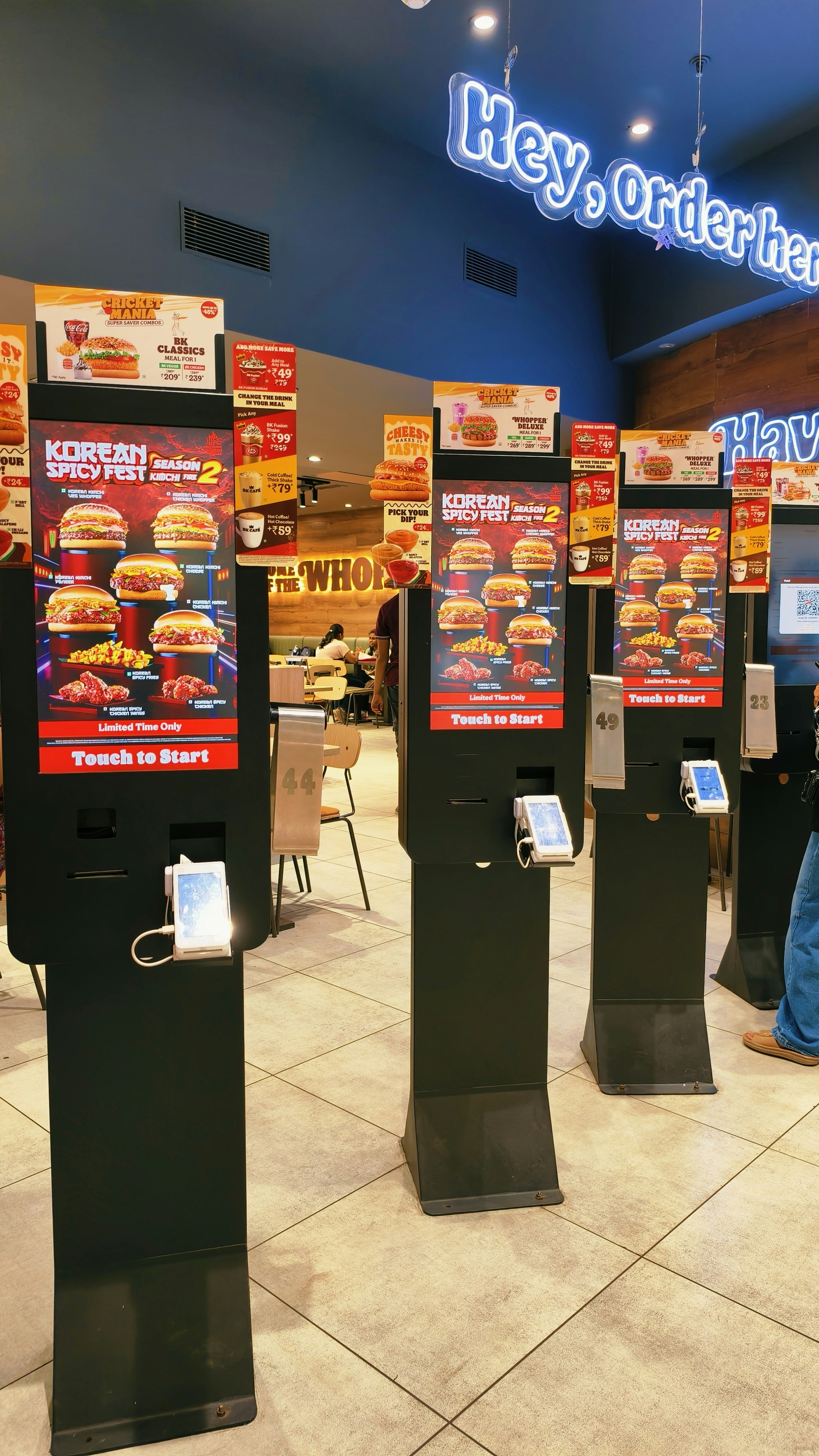 Energetic kiosk lineup inside a fast-food hall, featuring multiple bold posters promoting Korean Spicy Fest burgers. Clean tile floor, blue signage overhead, and patrons glimpsed in the background.