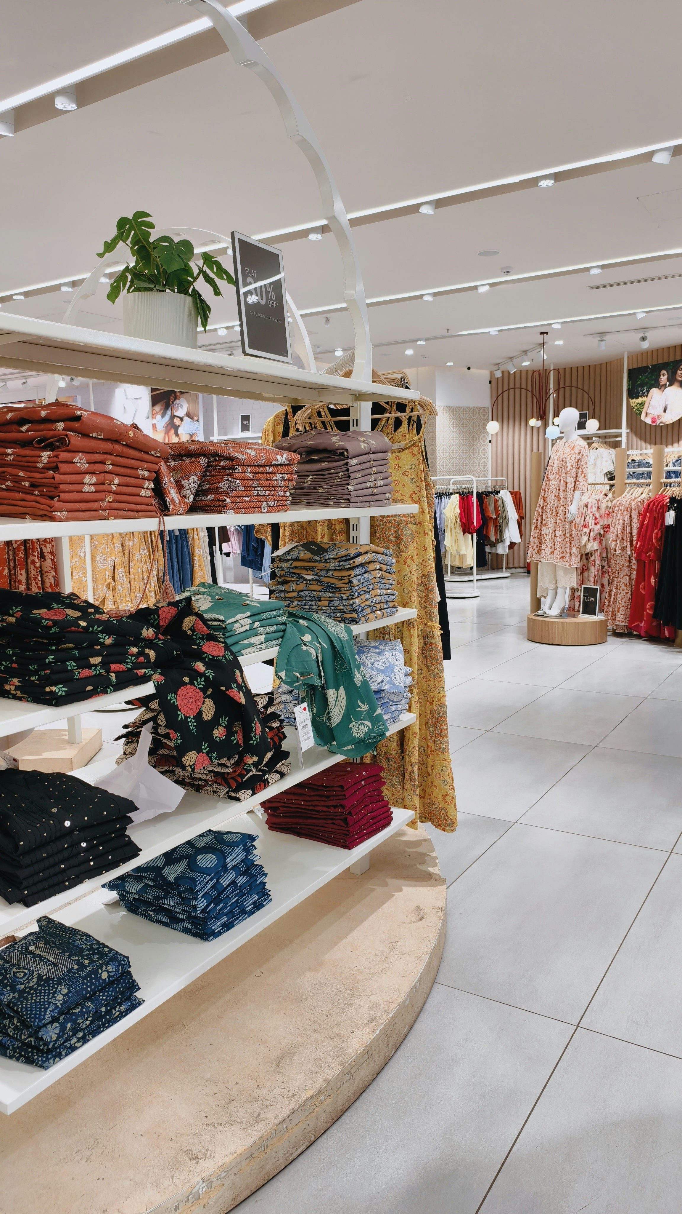 Rows of folded garments on white shelves in a modern retail interior with mannequins and hanging dresses in the background.