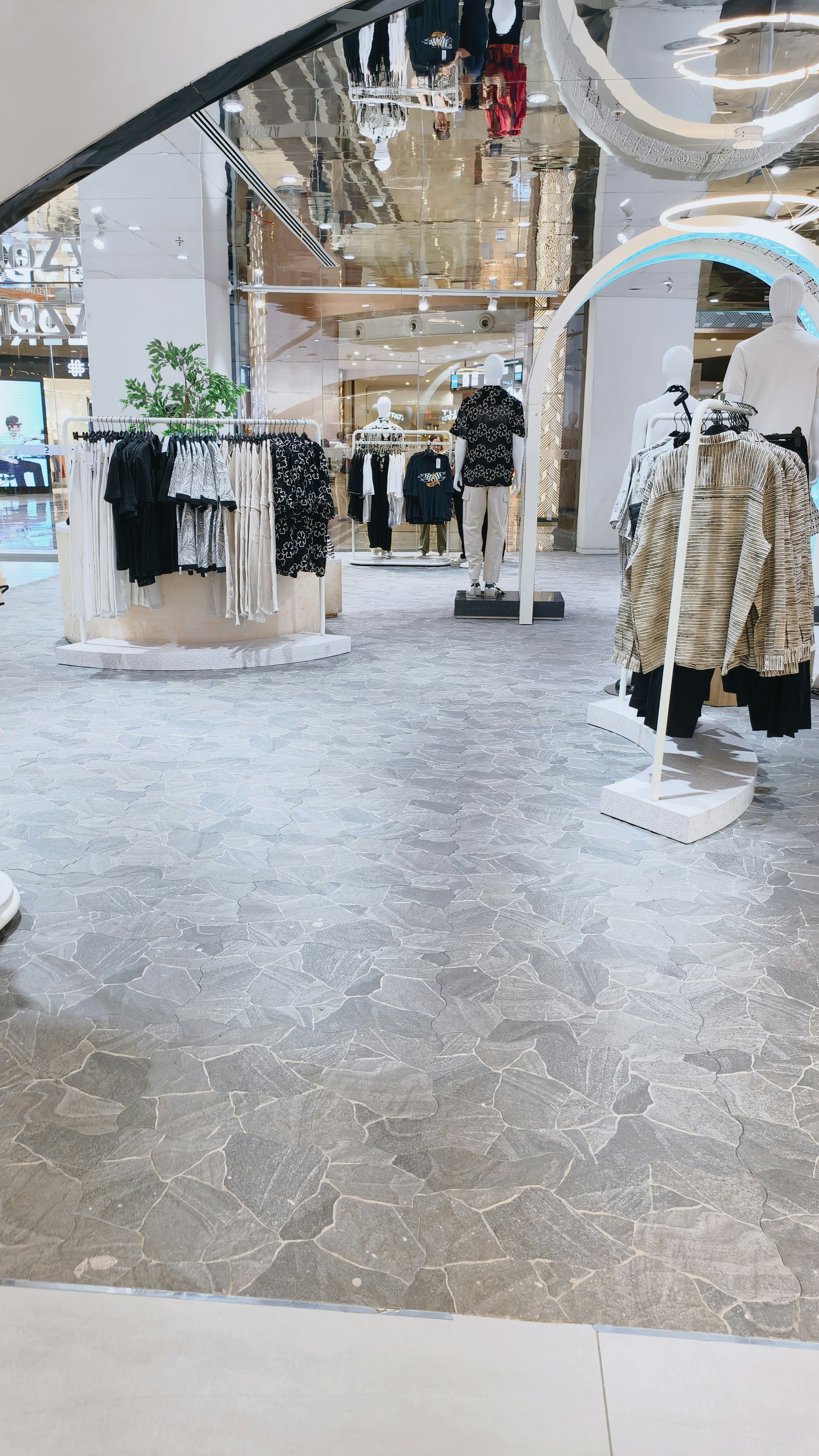 Clothing racks and mannequins arranged in a stylish boutique interior with tiled floor and reflective ceiling. A modern retail space highlighting apparel on circular stands and hanging racks.