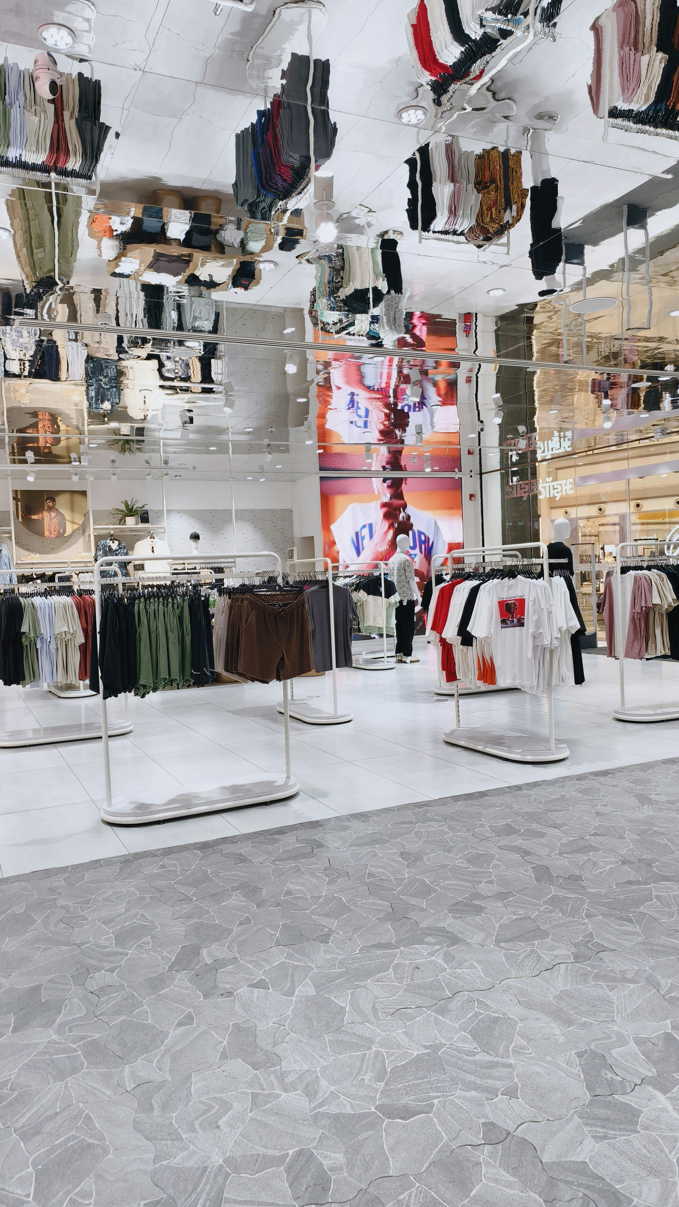 A brightly lit clothing store with racks of apparel and a highly reflective ceiling that creates a mirrored panorama. Colorful garments line the floor, while a large digital display dominates the back wall.