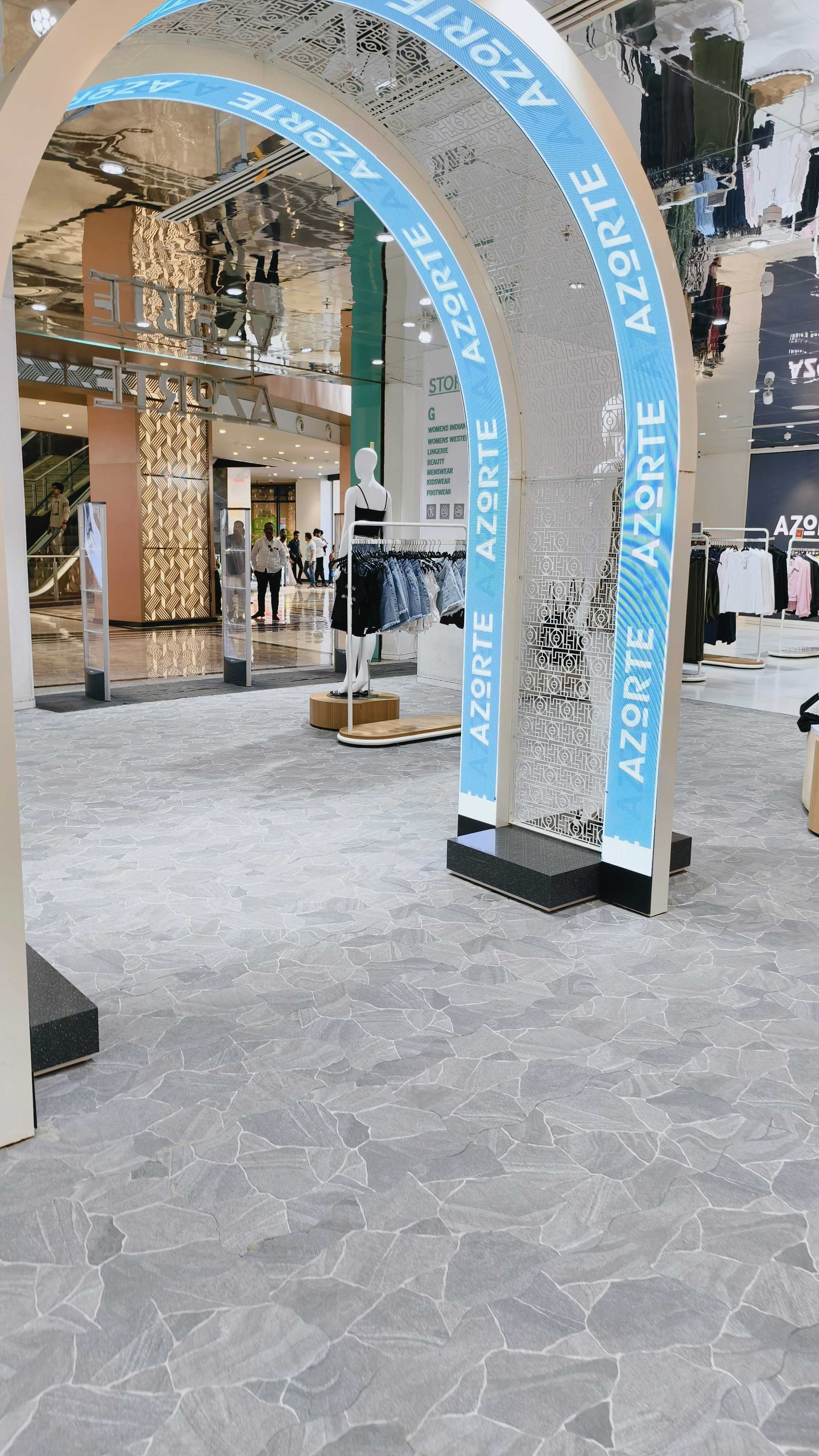 Bright boutique interior with curved blue archway poles labeled AZORTÉ framing a retail space and mannequins; polished stone floor and storefronts visible in the background.