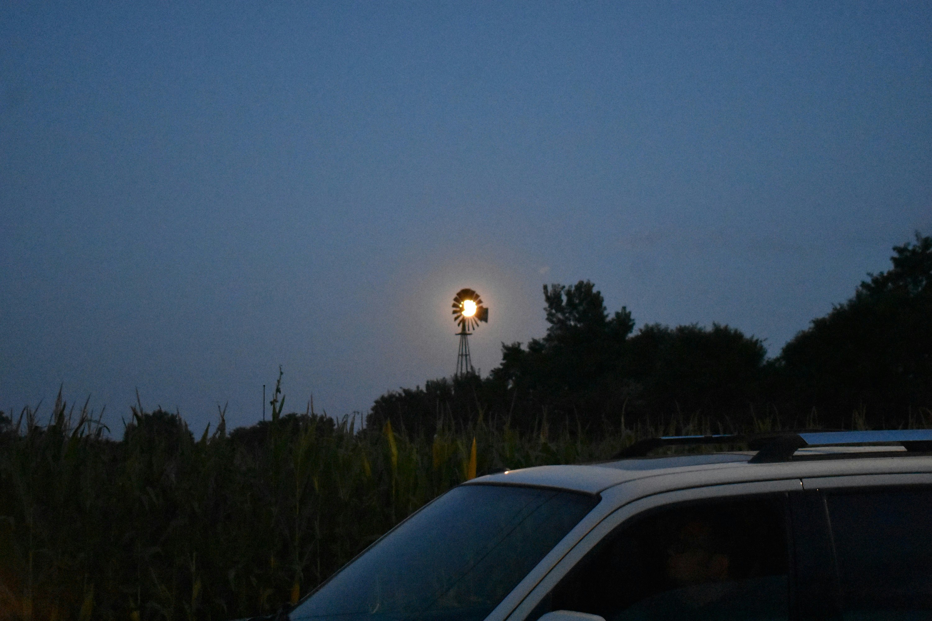 Dawn-blue sky with a roadside light tower glowing above tall grasses, a white SUV parked at the edge; a calm, humbling rural scene.