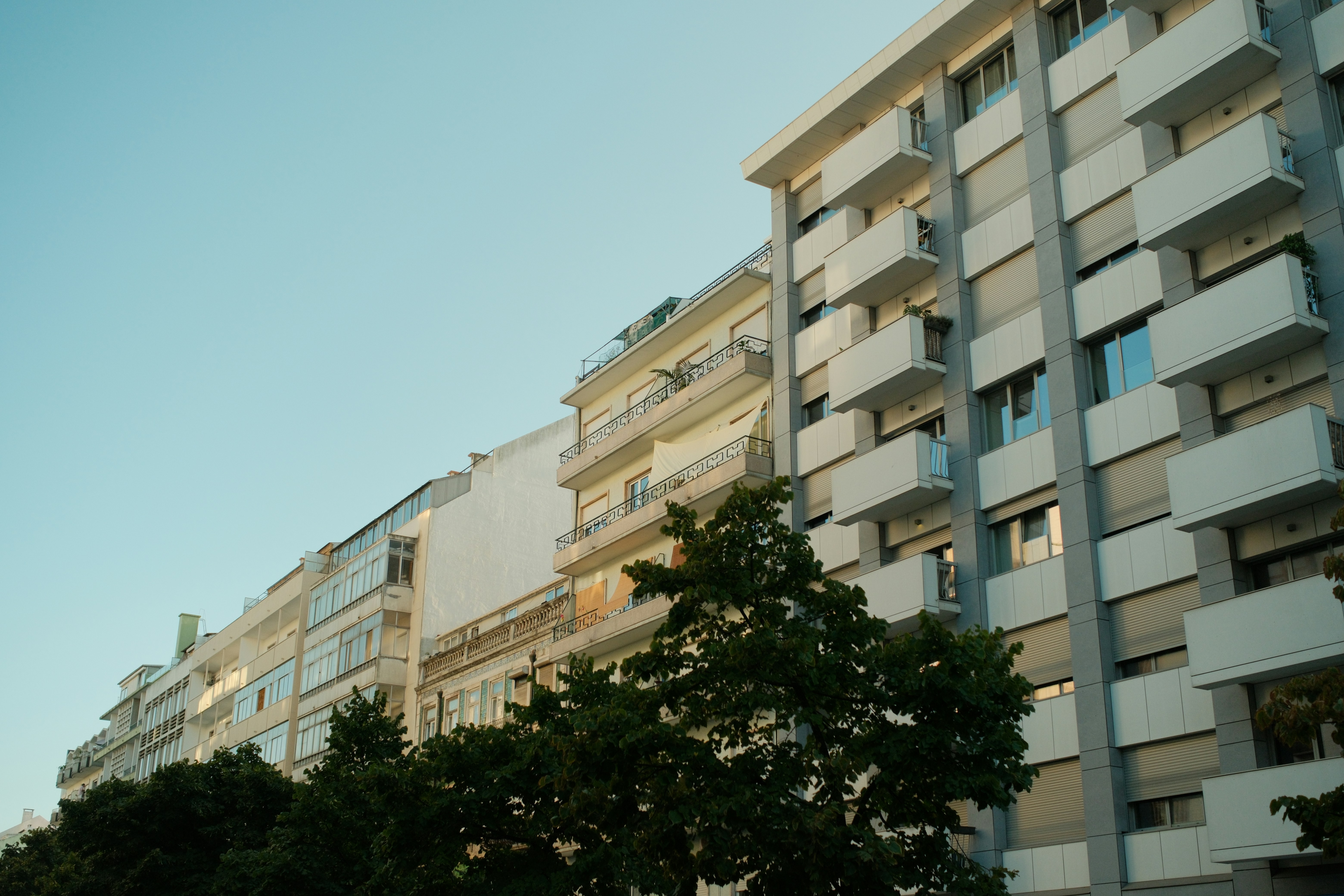 Layers of residential architecture from different periods rise above mature street trees along Avenida Almirante Reis in Lisbon, Portugal, under a clear summer sky in August 2026. Shot with Fujifilm X-M5 // Edifícios residenciais de diferentes épocas erguem-se sobre árvores maduras ao longo da Avenida Almirante Reis, em Lisboa, Portugal, sob um céu limpo de verão em agosto de 2026.