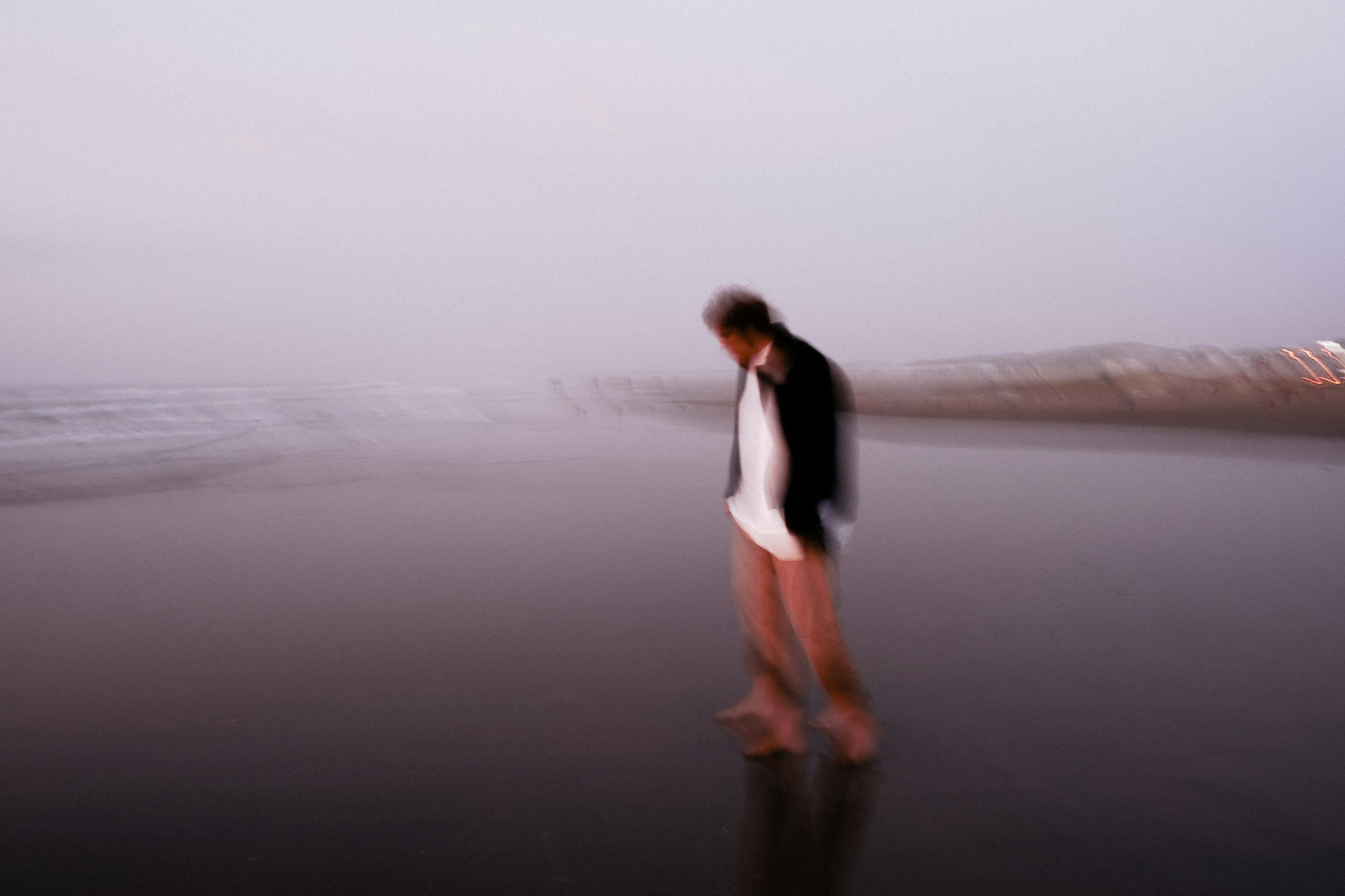 A lone, blurred figure stands near the water's edge on a sandy shore during blue hour; the scene emphasizes motion and mood with soft hues and a calm, expansive background.