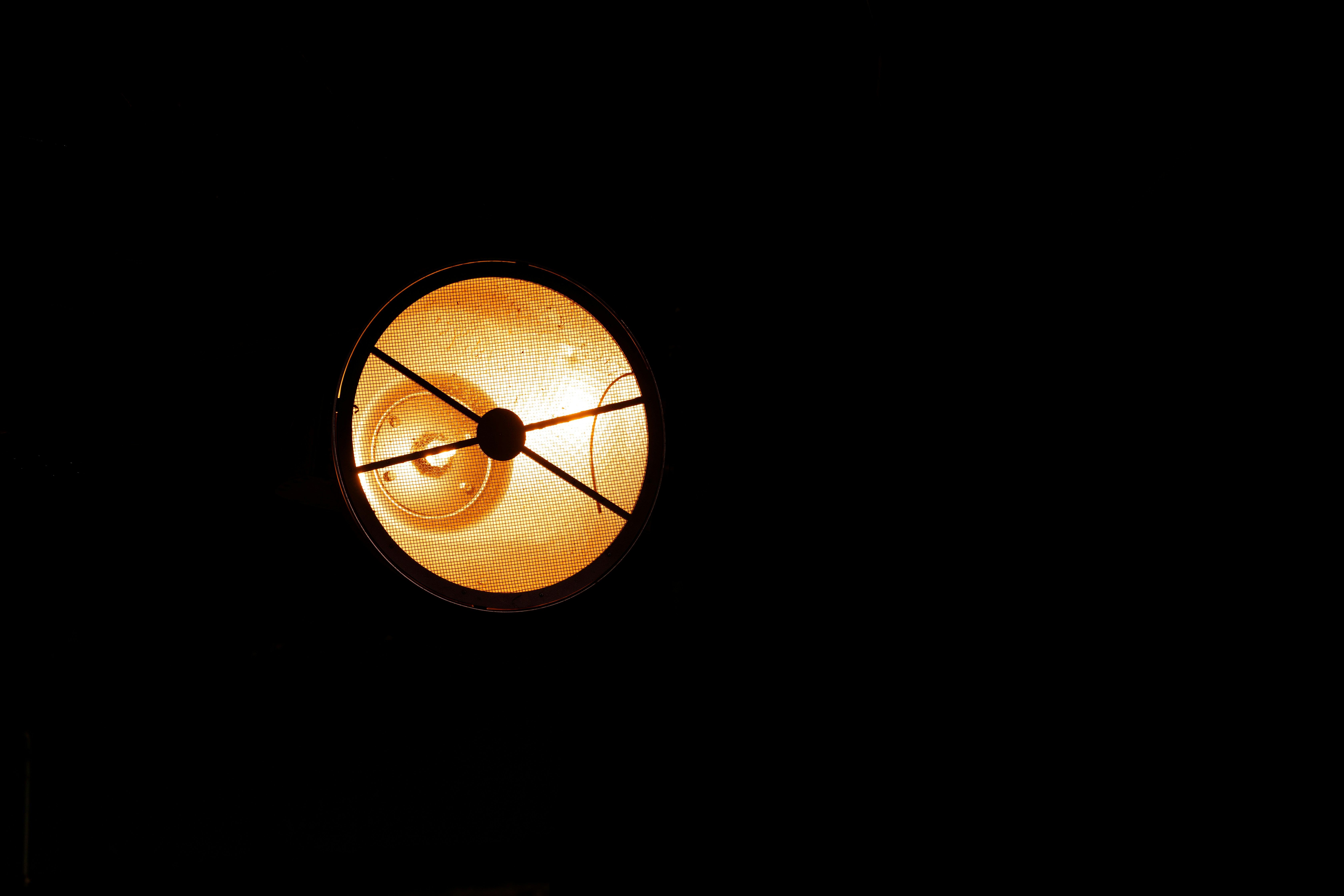 A warm, circular ceiling lamp glows softly in a dark space, revealing a metallic frame and glass with an amber hue. A minimal, isolated fixture that serves as the focal point in an otherwise void scene.