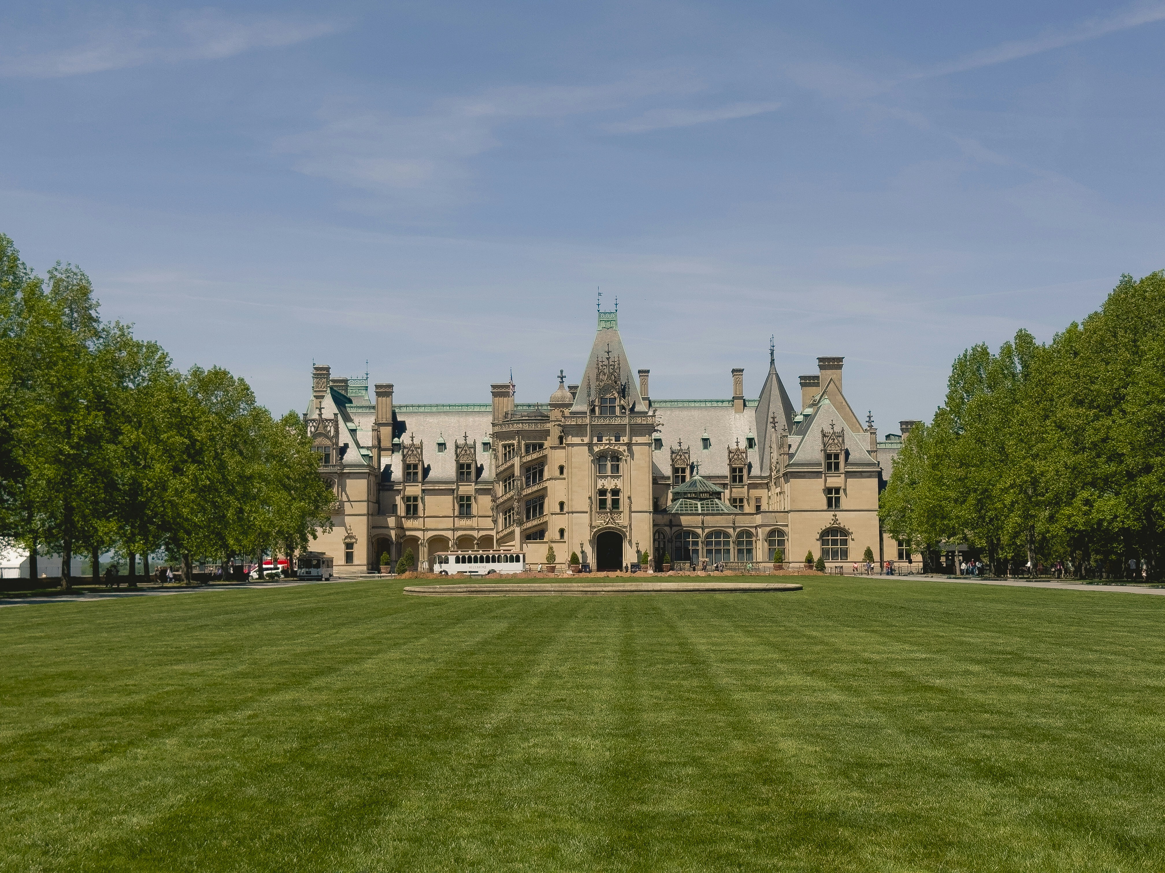 Grand university building with ornate stonework sits at the end of a wide, trimmed lawn and a central pathway; a bright blue sky and green trees enclose the scene. (Logo-like storefront not present; architectural exterior.)