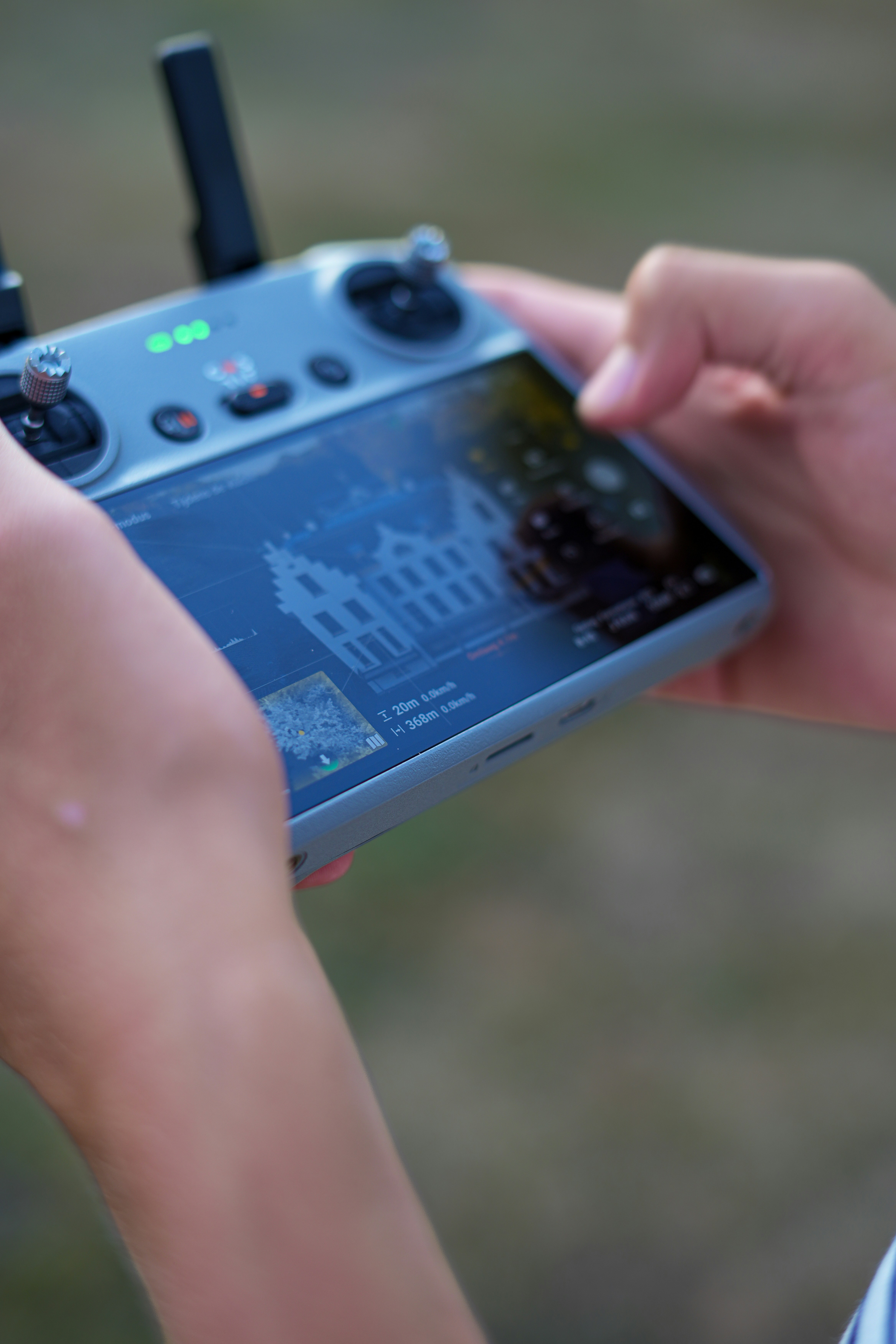 A handheld drone/remote controller screen shows a faint architectural display; hands grip the device as the screen glows with interface graphics. This is a close-up shot of an electronic control panel.