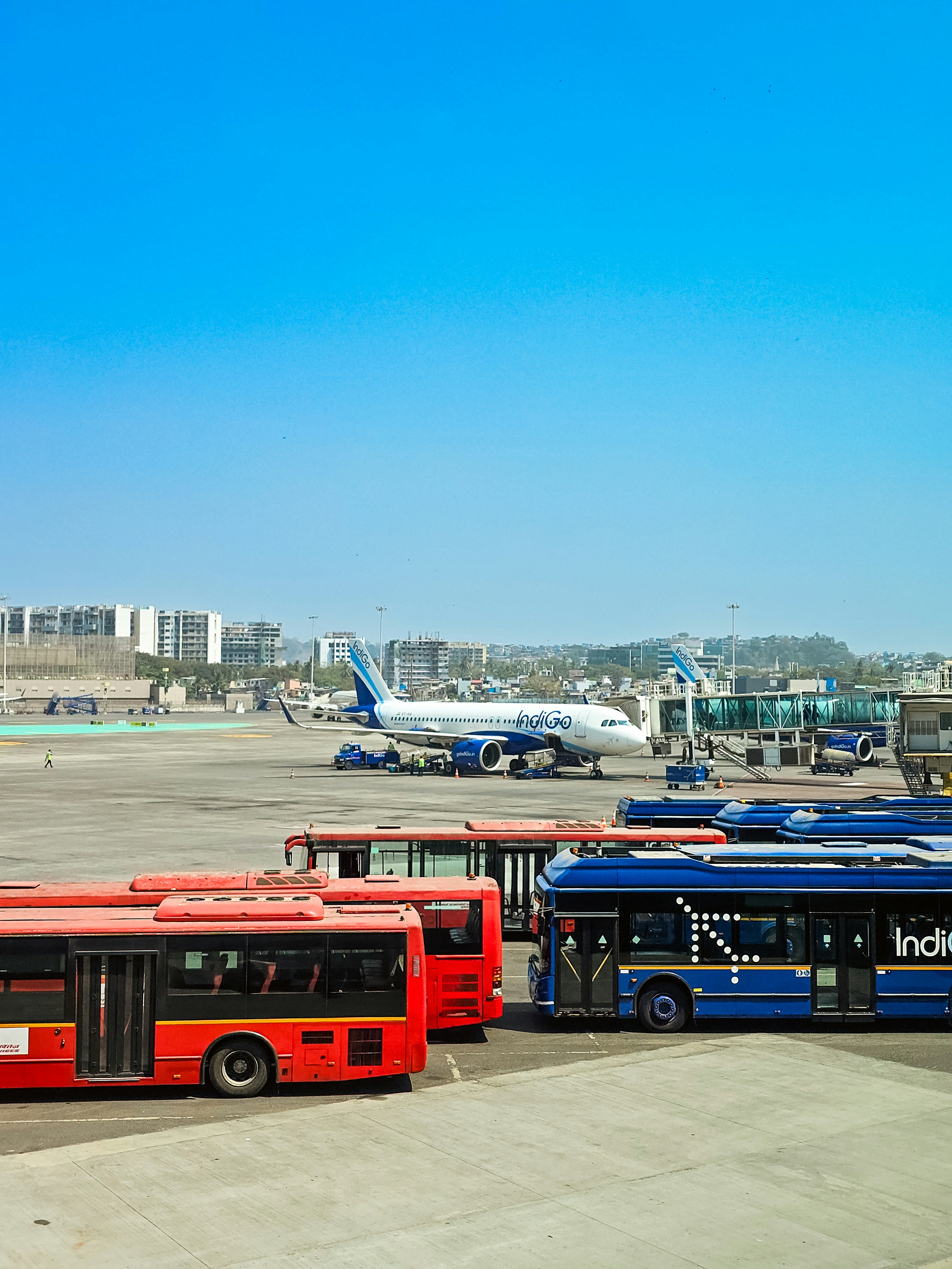 An IndiGo Airlines Airbus A320 271N narrow body passenger aircraft parked on the tarmac at Chhatrapati Shivaji Maharaj International Airport in Mumbai, India, connected to a terminal aerobridge. Several airport passenger buses, including red and blue buses, are parked in front of the aircraft and used to transport passengers between the terminal and aircraft at remote airport stands. The scene captures everyday ground operations at one of India's busiest airports.