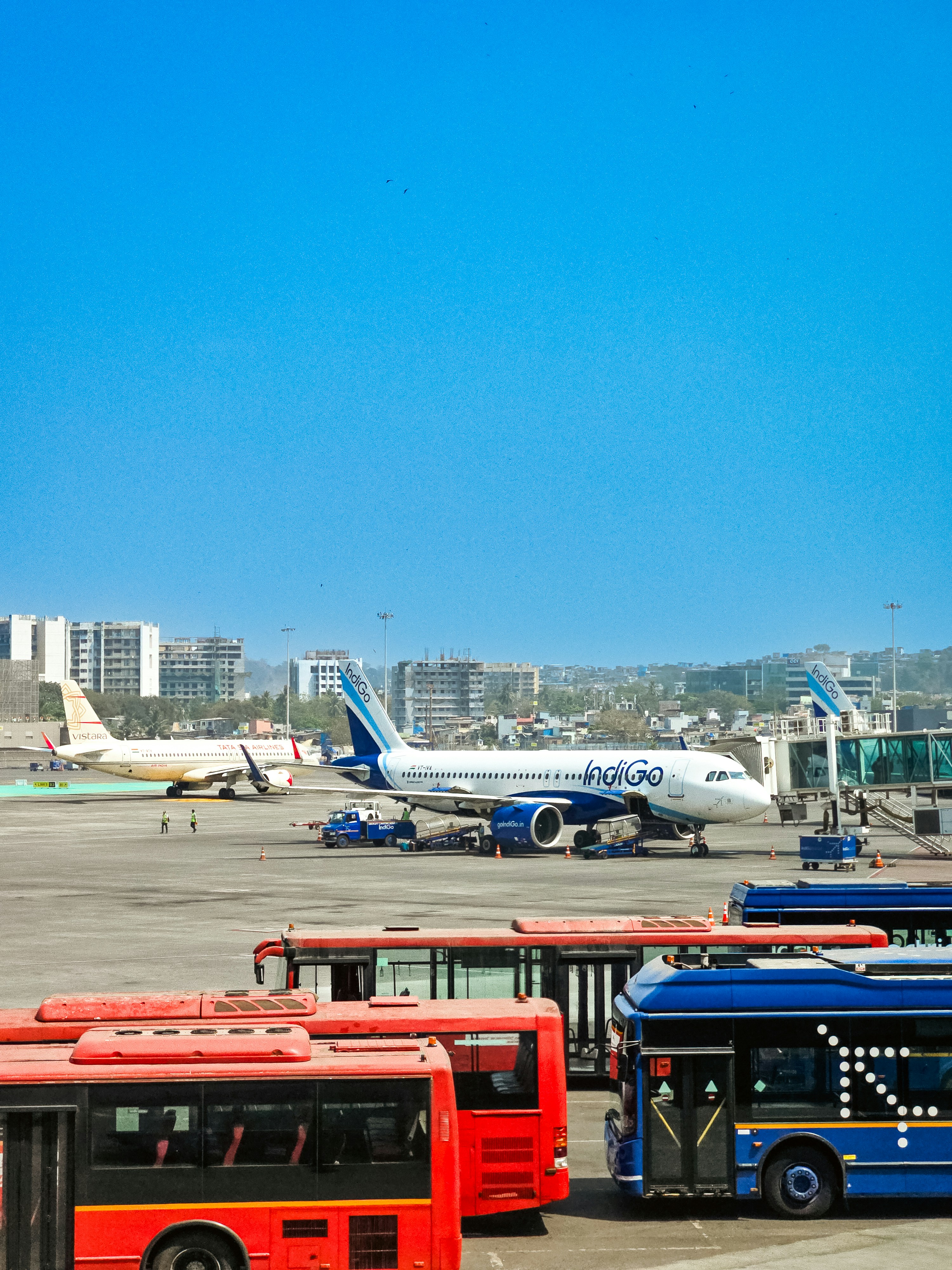 An IndiGo Airlines Airbus A320 271N narrow body passenger aircraft parked on the tarmac at Chhatrapati Shivaji Maharaj International Airport in Mumbai, India, connected to a terminal aerobridge. Several airport passenger buses, including red and blue buses, are parked in front of the aircraft and used to transport passengers between the terminal and aircraft at remote airport stands. The scene captures everyday ground operations at one of India's busiest airports.