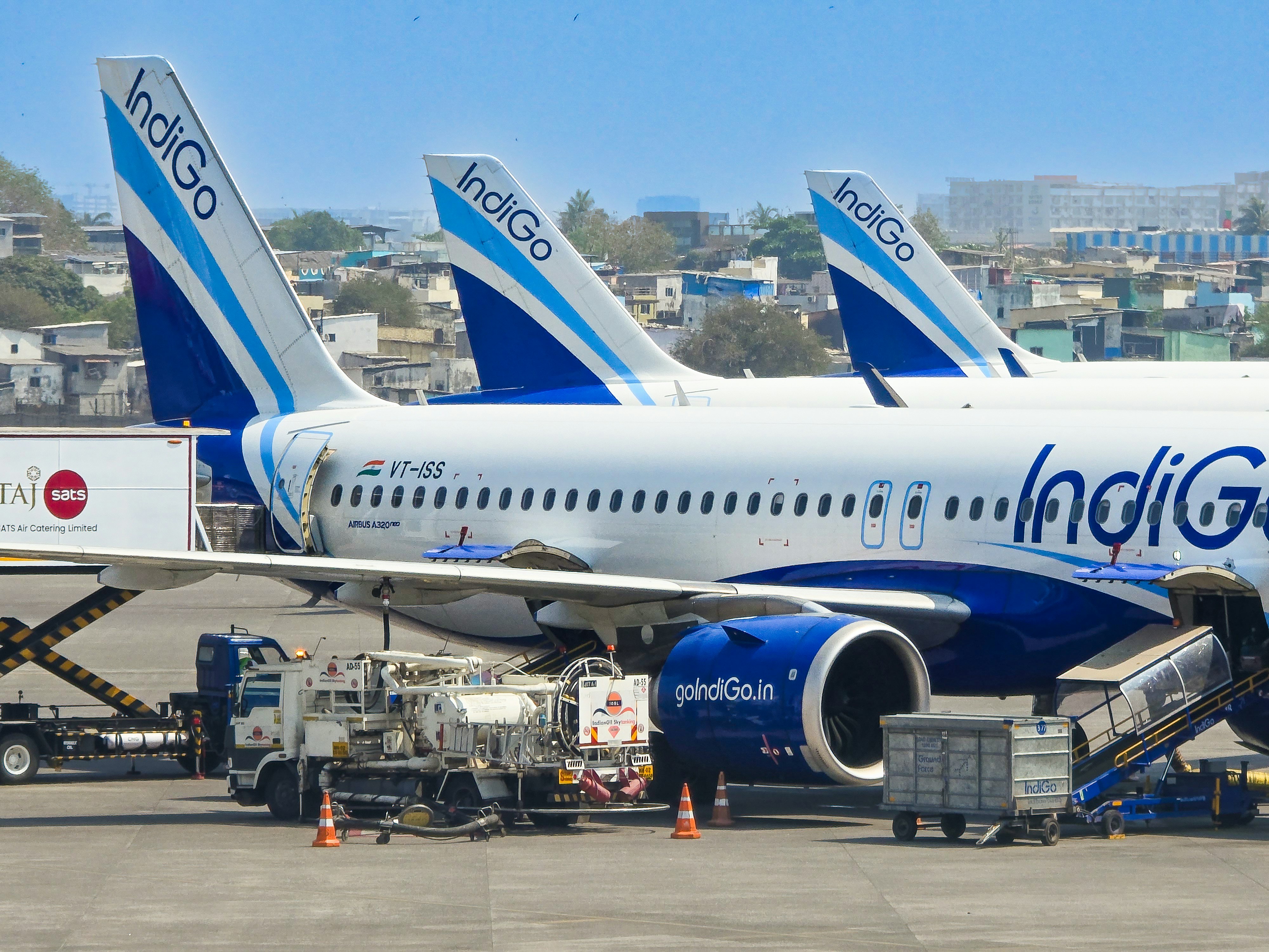 An IndiGo Airbus A320 251N narrow body passenger aircraft parked at Chhatrapati Shivaji Maharaj International Airport in Mumbai, India. The aircraft is positioned at a terminal gate with an airbridge connected to its forward door, while the tails of two additional IndiGo aircraft parked nearby are visible in the background. The scene captures the busy operations and distinctive blue and white livery of India's largest airline at Mumbai airport.