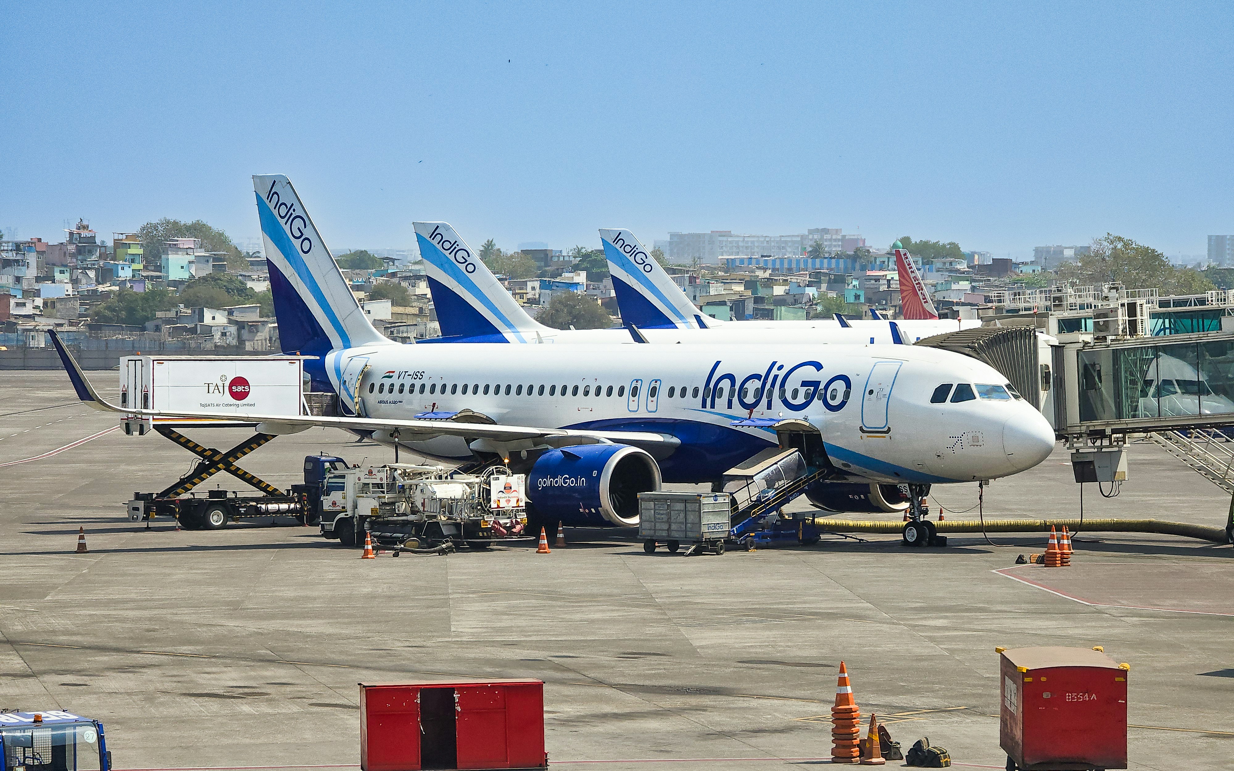 An IndiGo Airbus A320 251N narrow body passenger aircraft parked at Chhatrapati Shivaji Maharaj International Airport in Mumbai, India. The aircraft is positioned at a terminal gate with an airbridge connected to its forward door, while the tails of two additional IndiGo aircraft parked nearby are visible in the background. The scene captures the busy operations and distinctive blue and white livery of India's largest airline at Mumbai airport.