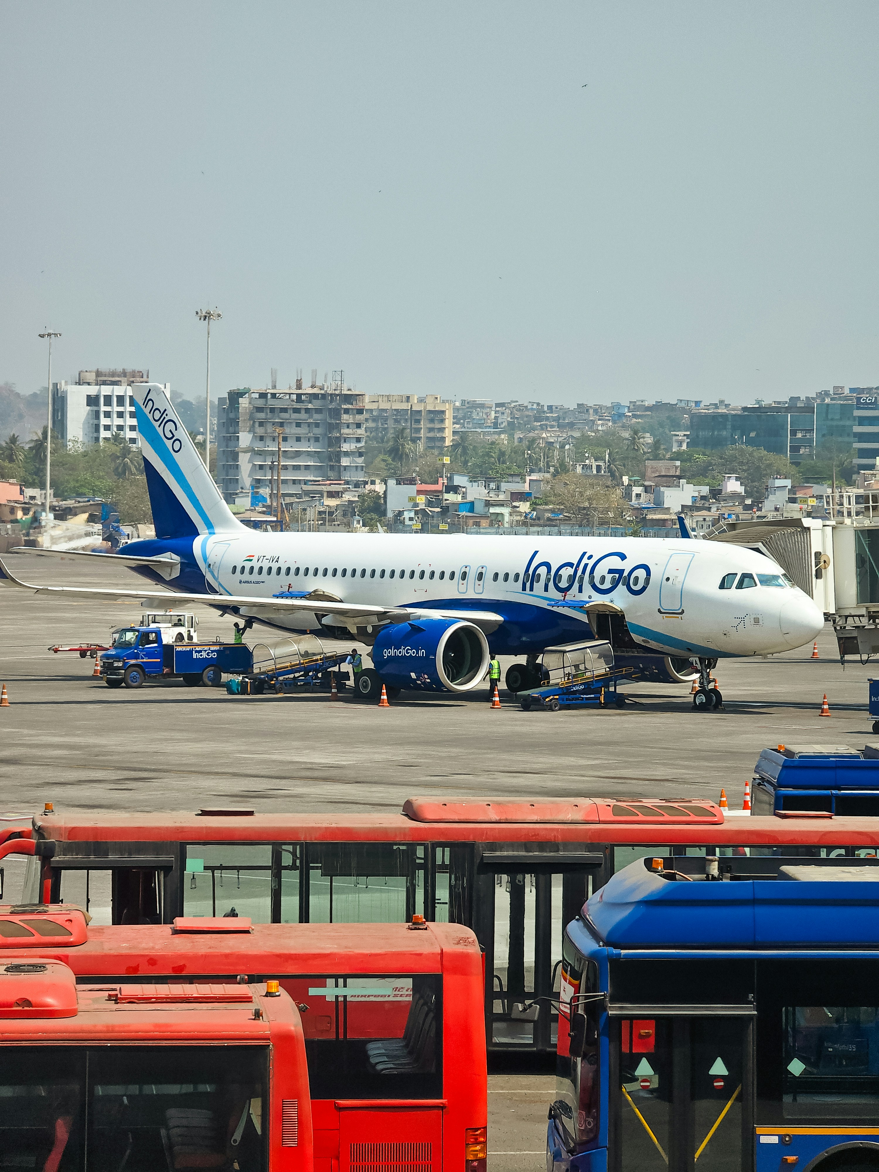 An IndiGo Airlines Airbus A320 271N narrow body passenger aircraft parked on the tarmac at Chhatrapati Shivaji Maharaj International Airport in Mumbai, India, connected to a terminal aerobridge. Several airport passenger buses, including red and blue buses, are parked in front of the aircraft and used to transport passengers between the terminal and aircraft at remote airport stands. The scene captures everyday ground operations at one of India's busiest airports.