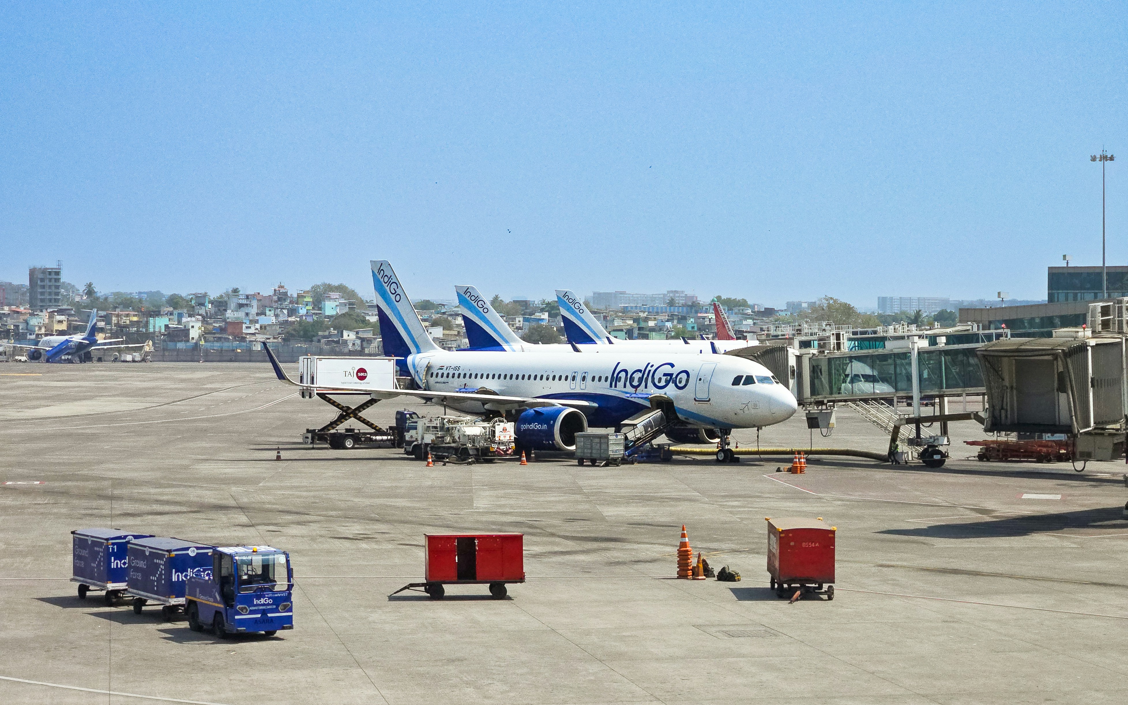 An IndiGo Airbus A320 251N narrow body passenger aircraft parked at Chhatrapati Shivaji Maharaj International Airport in Mumbai, India. The aircraft is positioned at a terminal gate with an airbridge connected to its forward door, while the tails of two additional IndiGo aircraft parked nearby are visible in the background. The scene captures the busy operations and distinctive blue and white livery of India's largest airline at Mumbai airport.