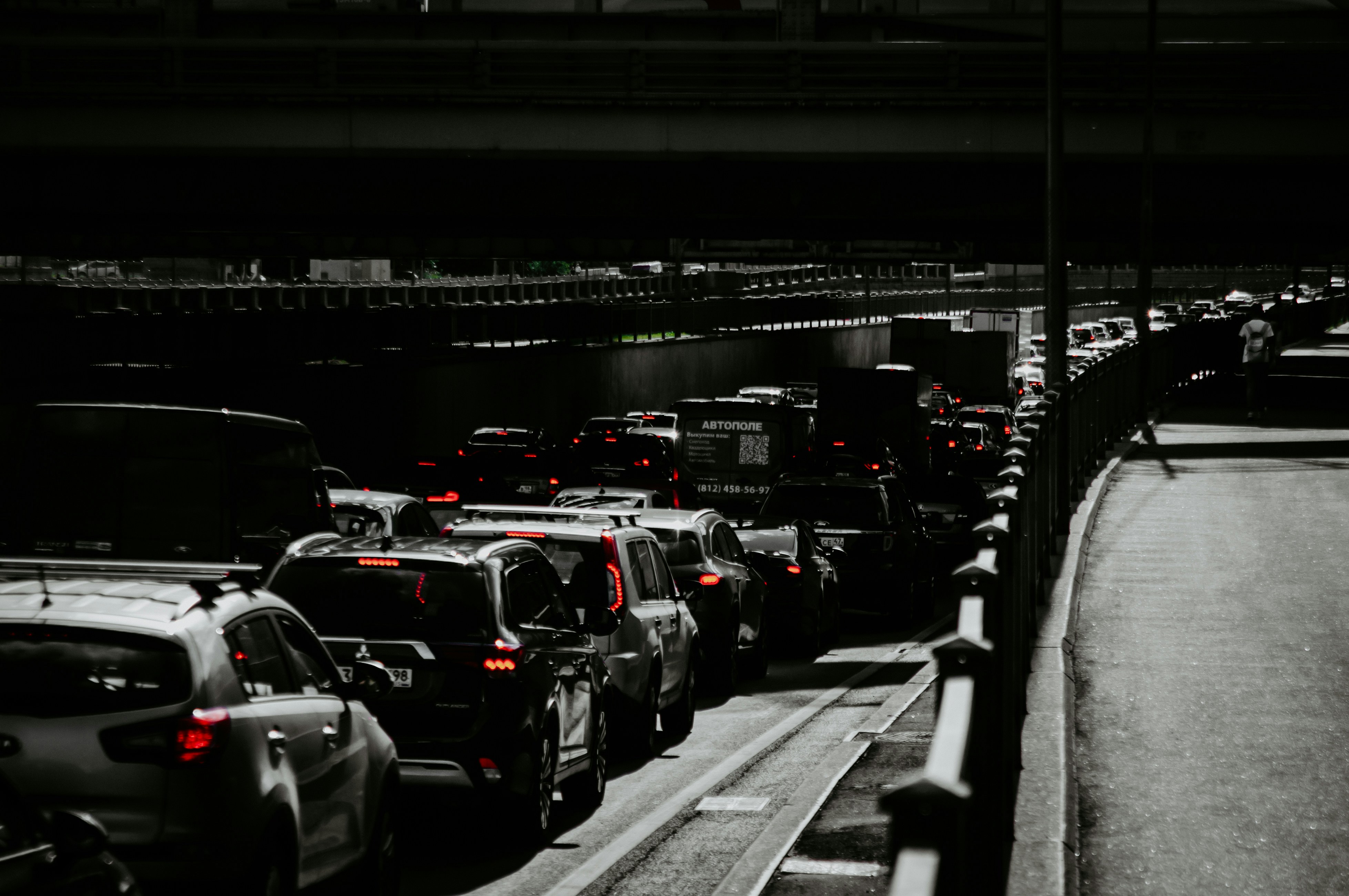 Dense traffic compresses into a narrow underpass, with taillights forming a crimson line along the lane. A sunlit barrier edge hints at the roadside environment, while the upper structure frames the scene in a grayscale urban mood.