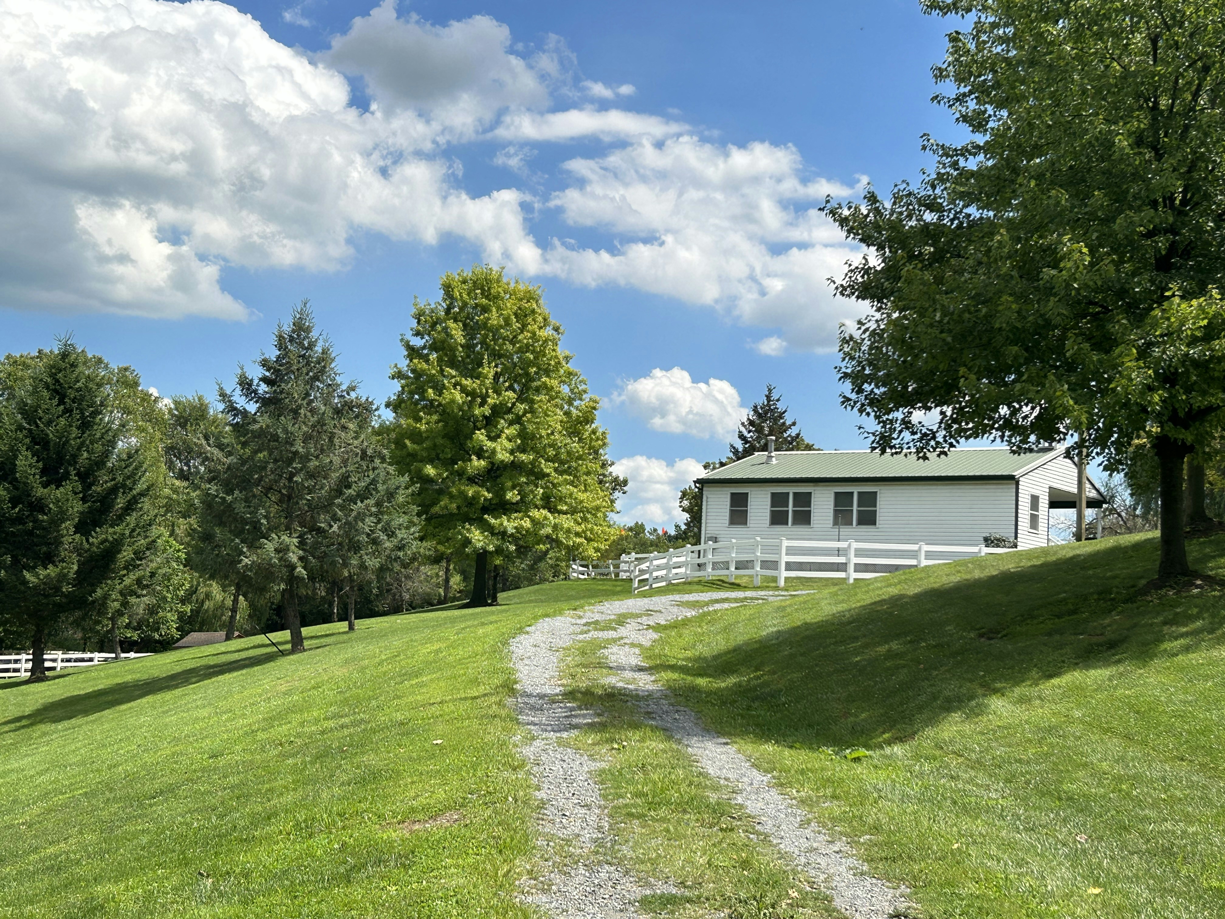 A gravel path winds up a grassy hill toward a white barn under a bright blue sky with fluffy clouds, flanked by trees.