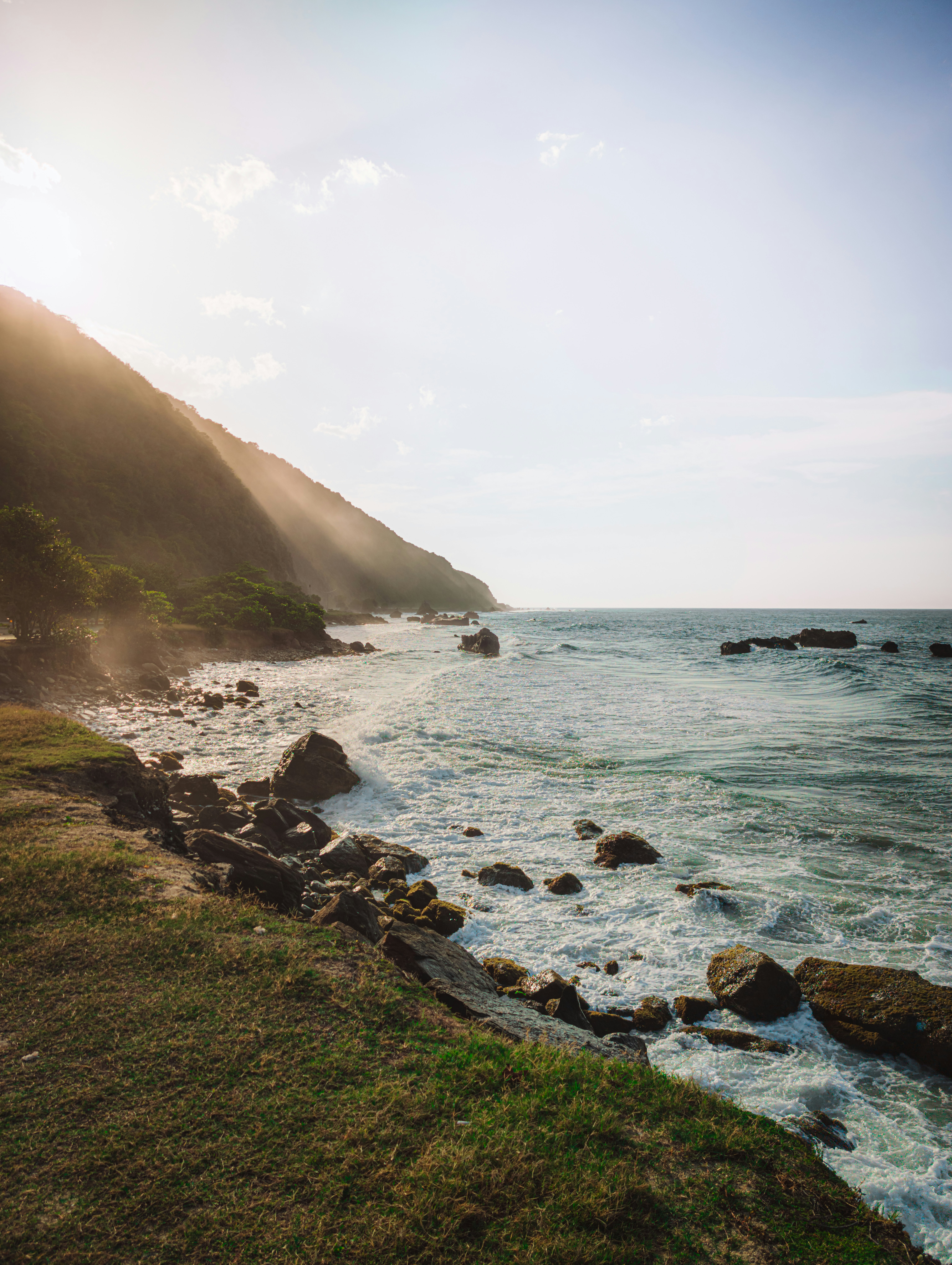 Coastal scene with rocky shoreline, gentle waves, and sunlit hillside on the left; serene horizon over calm sea.