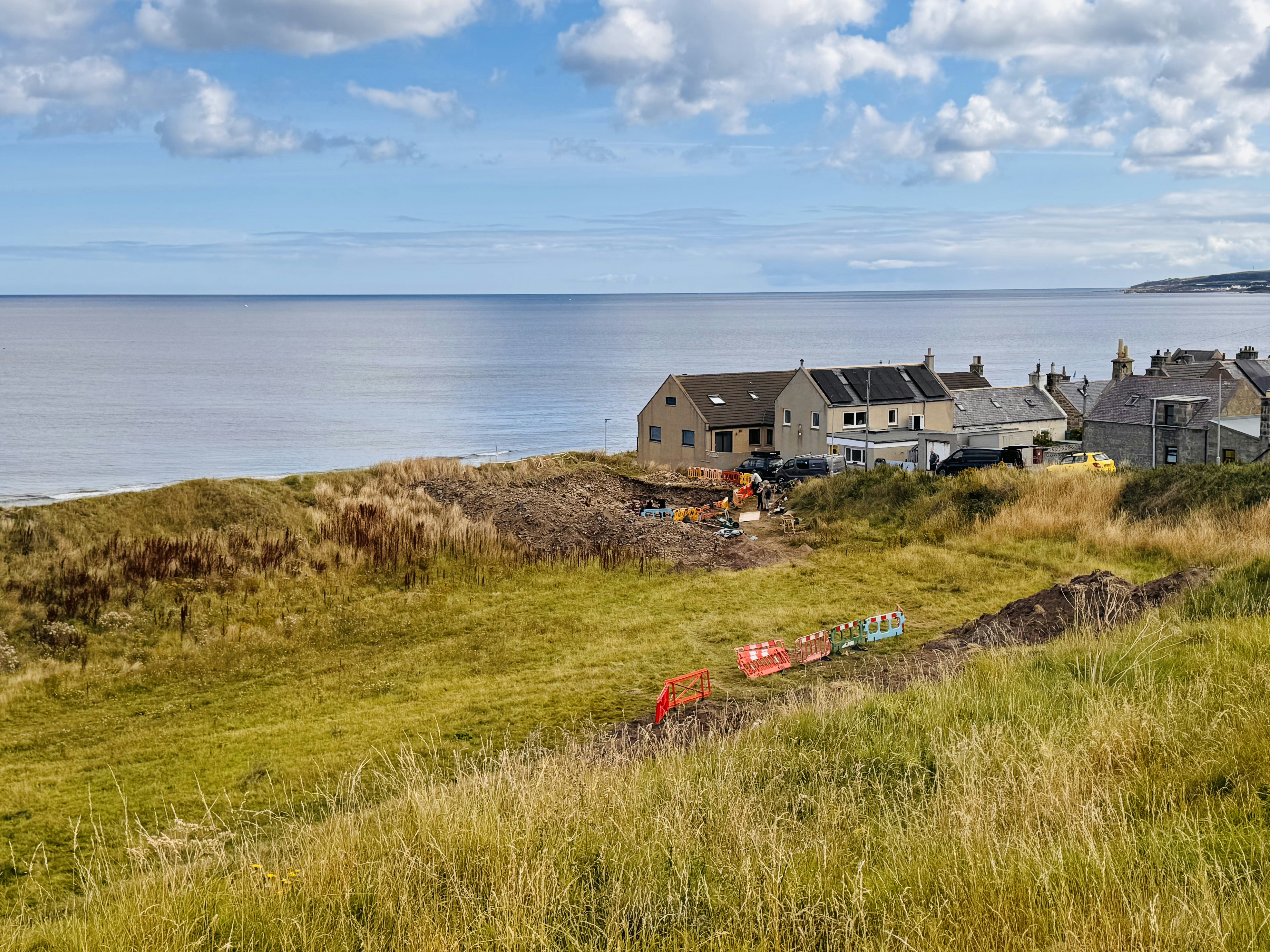 Coastal cottages sit above grassy dunes with a calm sea beyond; construction barriers and equipment hint at ongoing work near the shoreline.
