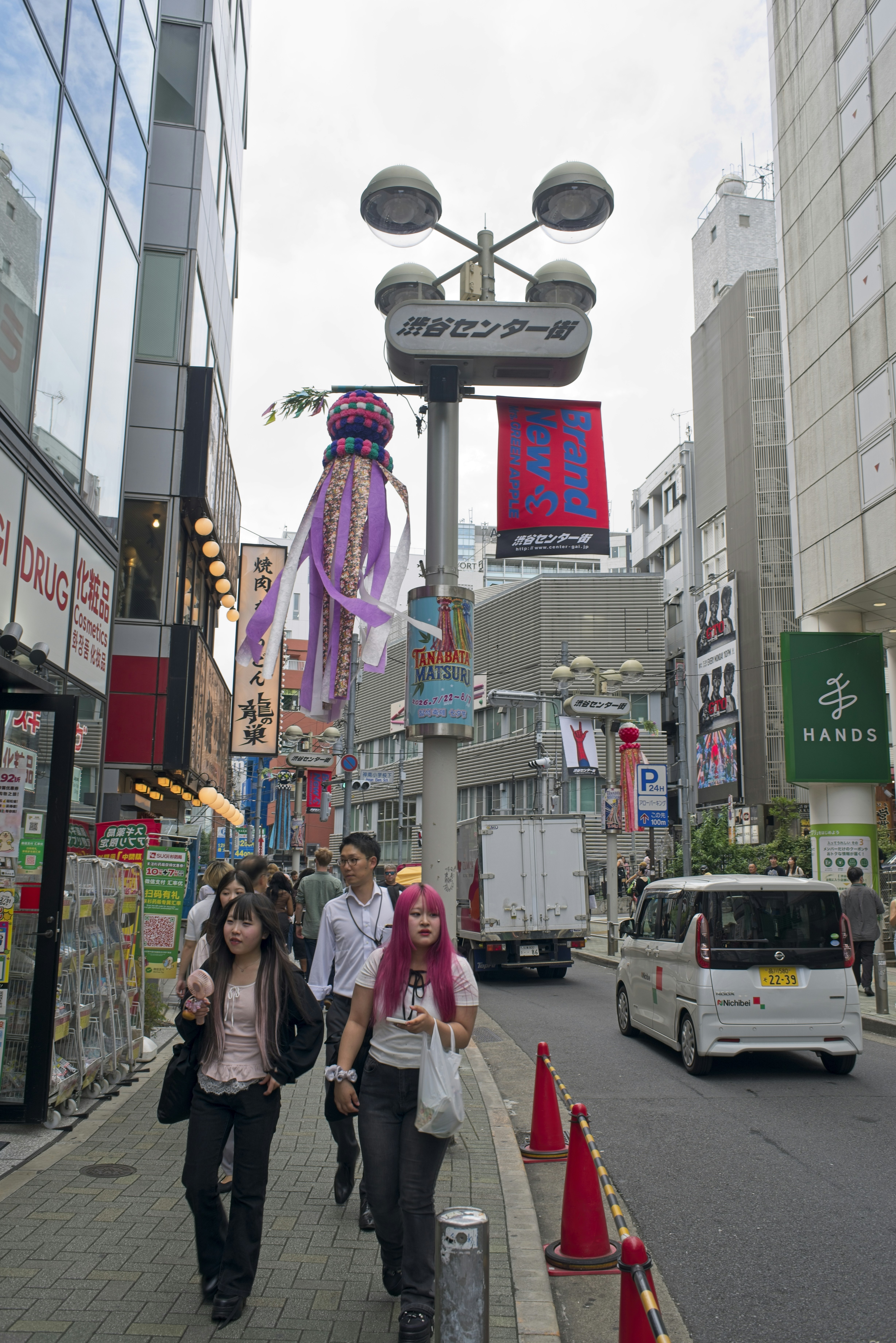 Pedestrians with vibrant pink hair and casual street style walk beneath a decorative streetlamp adorned with Tanabata Matsuri festival streamers in Shibuya Center Gai. Bold banners advertise "Brand New 3" and the summer festival dates, while nearby storefronts like HANDS and various Japanese shops line the busy street. Traffic cones and a delivery truck occupy the roadway as cars pass through the intersection. The scene captures Shibuya's blend of festive decoration, youthful fashion, and everyday urban movement on a warm July afternoon.