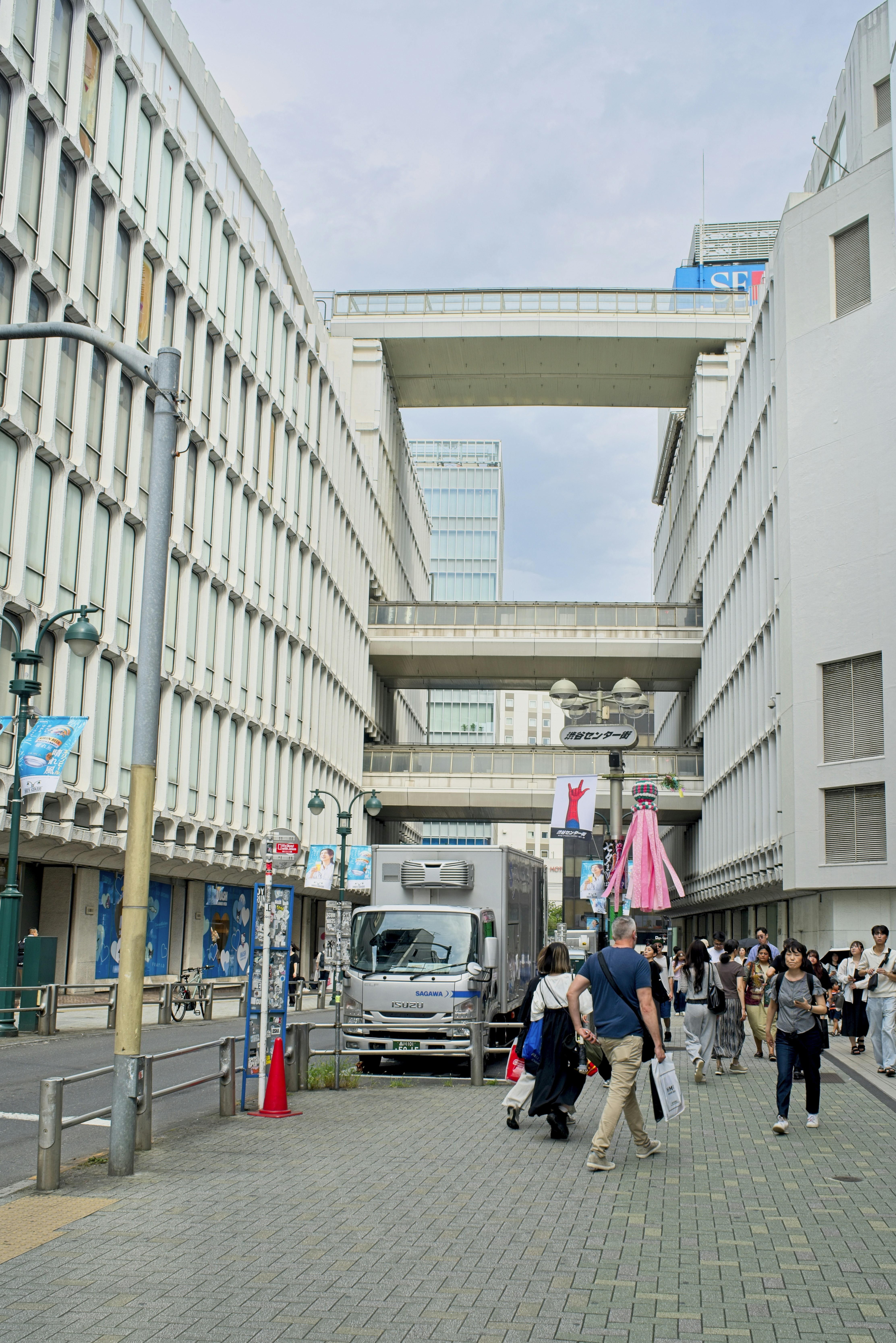 A series of stacked elevated walkways span between two symmetrical Shibuya buildings, creating a striking architectural corridor above a busy pedestrian street. A delivery truck sits parked below while pedestrians move through the paved plaza, passing beneath colorful Tanabata streamers hanging from a lamppost. The layered concrete overpasses and geometric building facades give the scene a distinctly urban, structured feel, capturing Shibuya's blend of pedestrian life and dense architectural infrastructure under a hazy summer evening sky.