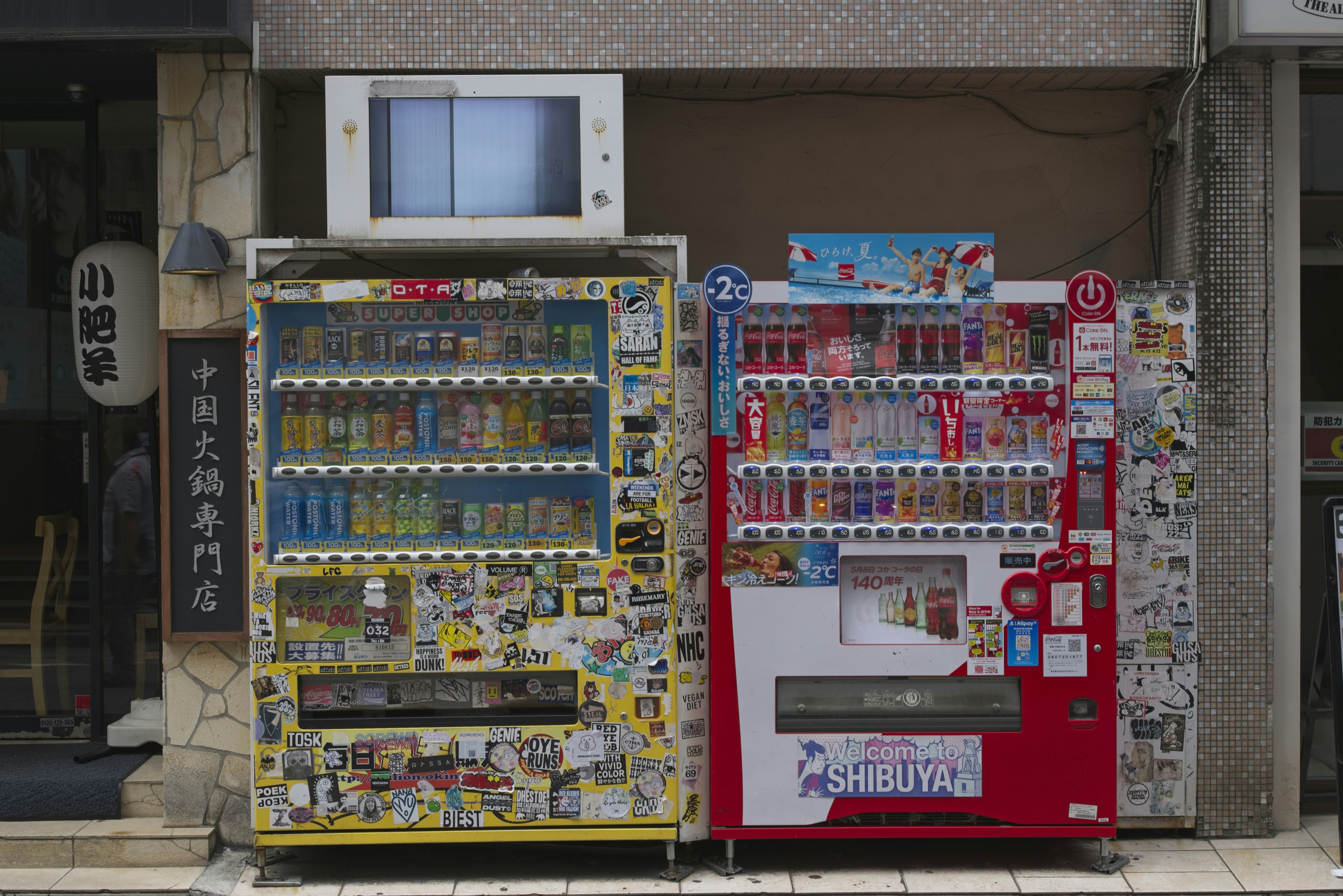 Two Sticker-Covered Vending Machines Guard a Busy Tokyo Street