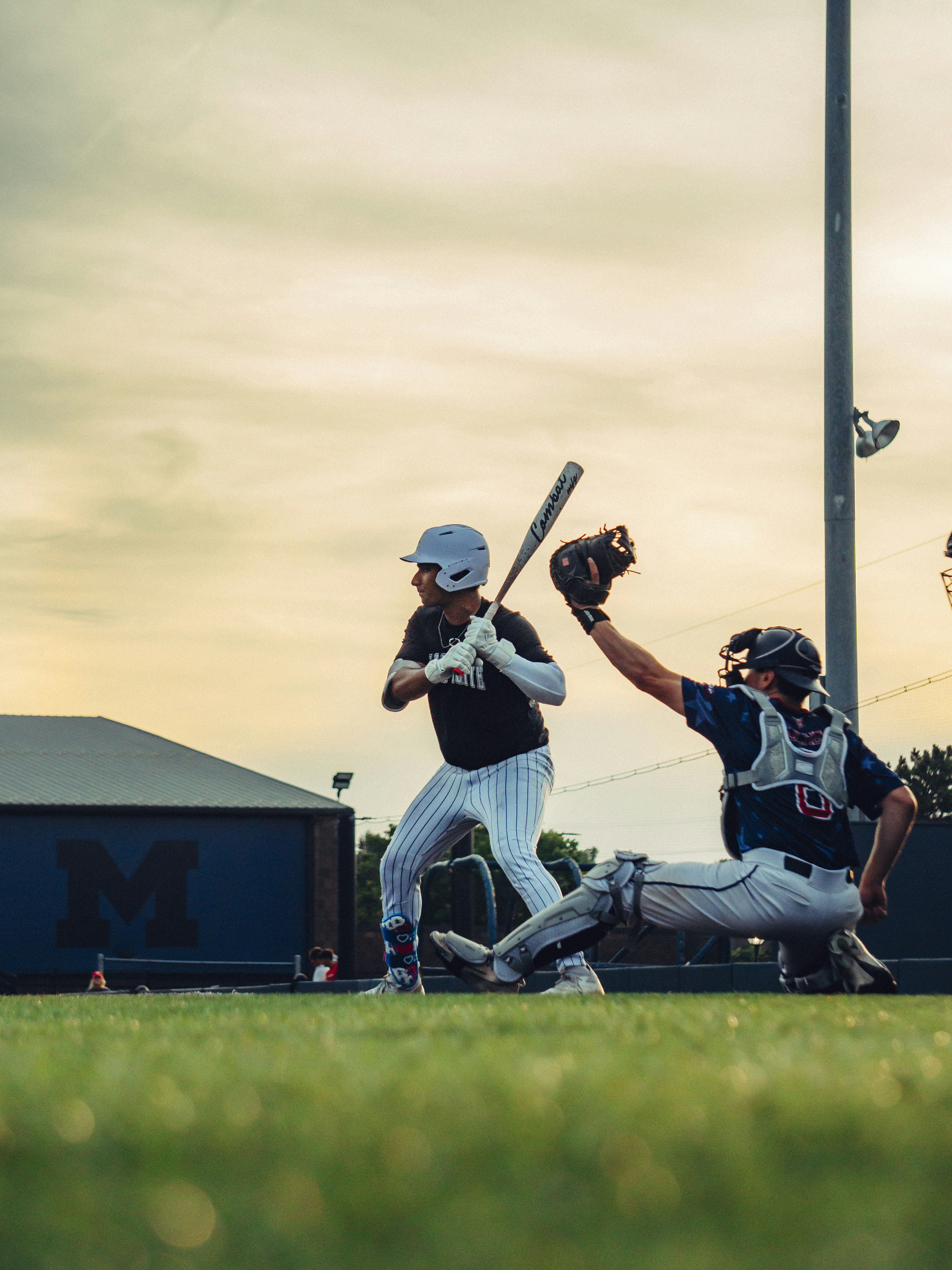 Swinging toward the on-deck glow: a batter and catcher in dusk-lit action