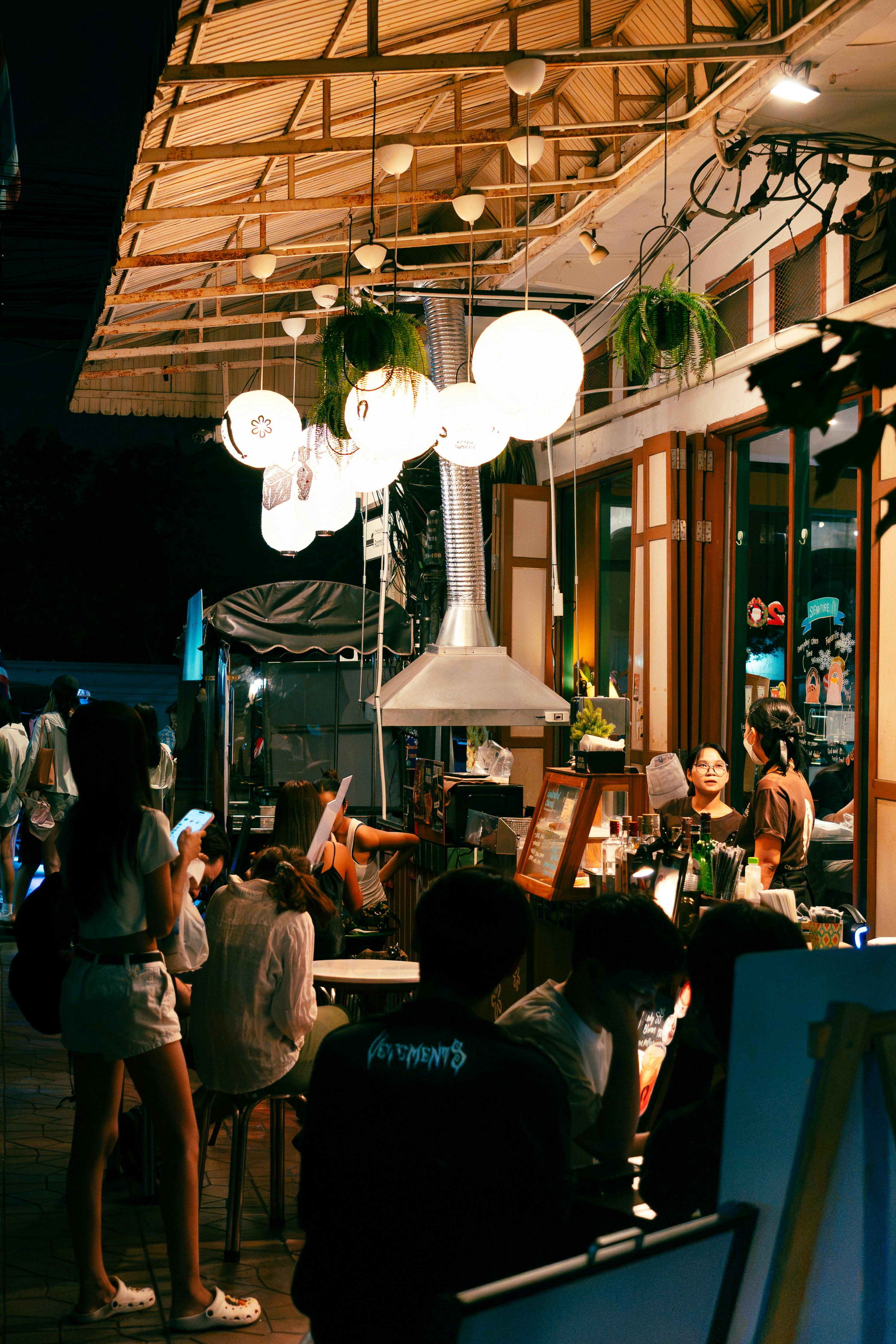 Ambient scene of a busy night market café with warm hanging lanterns, a centralized vent hood, and patrons seated around a counter as staff chat in the background.