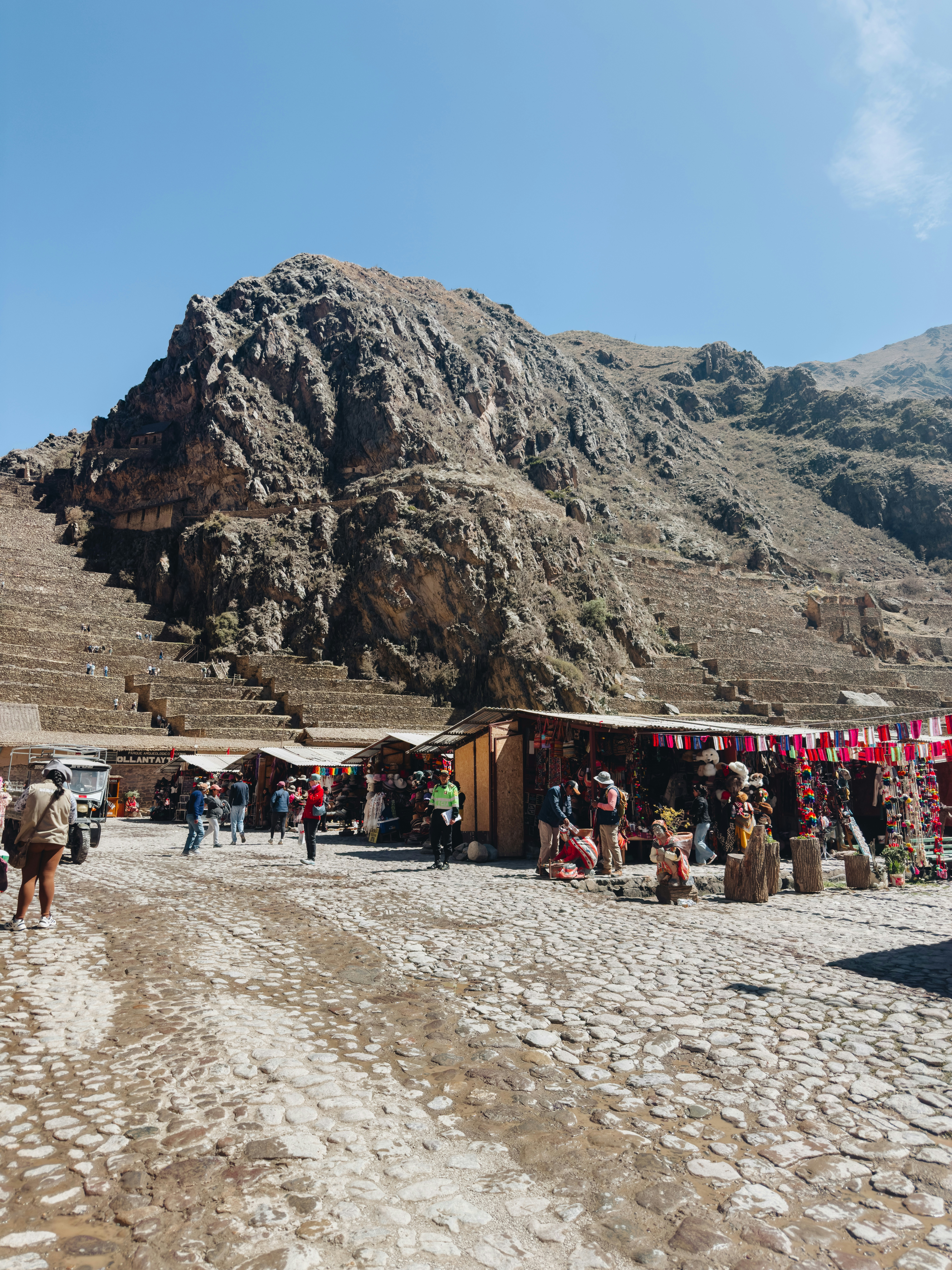 Stone amphitheater steps rise behind a bustling marketplace with colorful textiles and visitors; foreground cobblestones lead to small stalls.