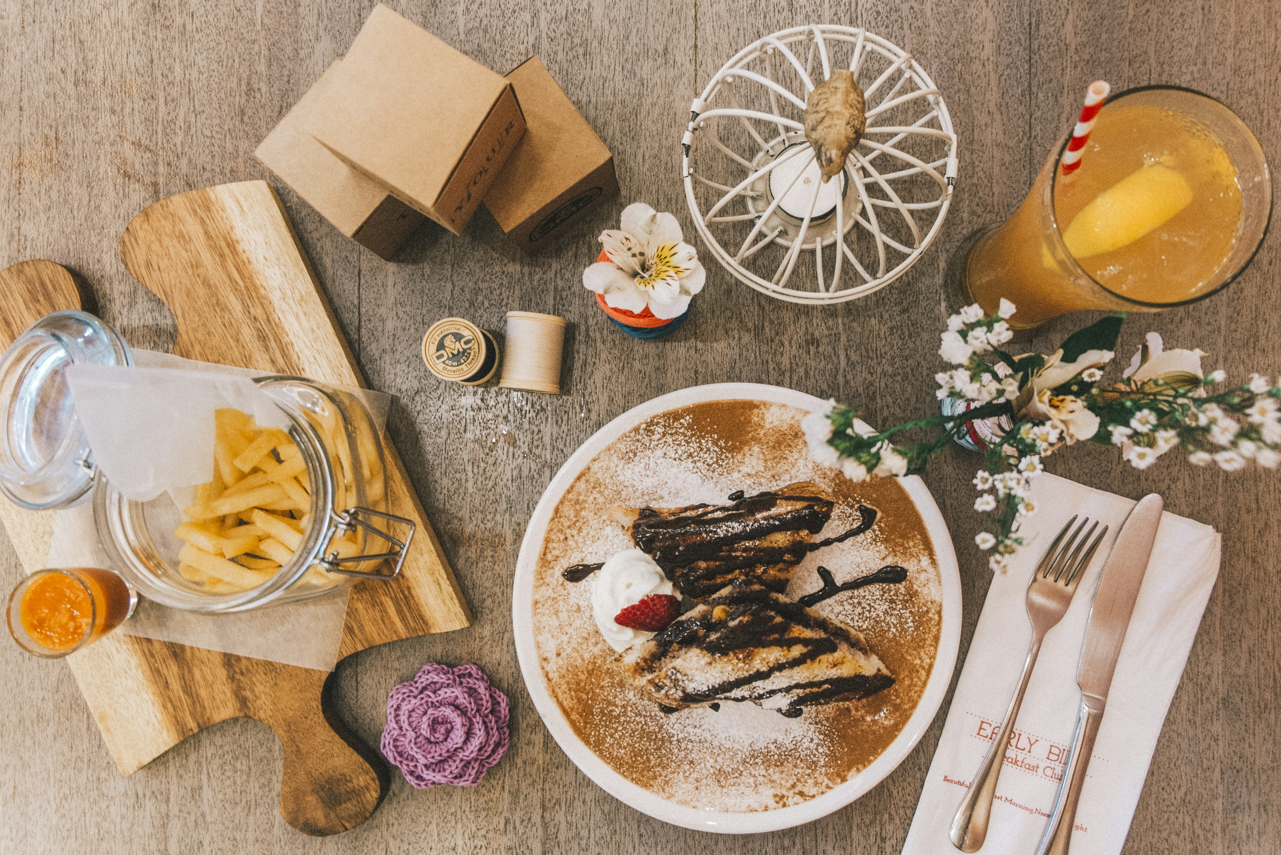 Top-down view of a dessert and beverage setup with a caramel-drizzled pancake, french fries, a glass jug, and seasonal decor on a wooden surface.